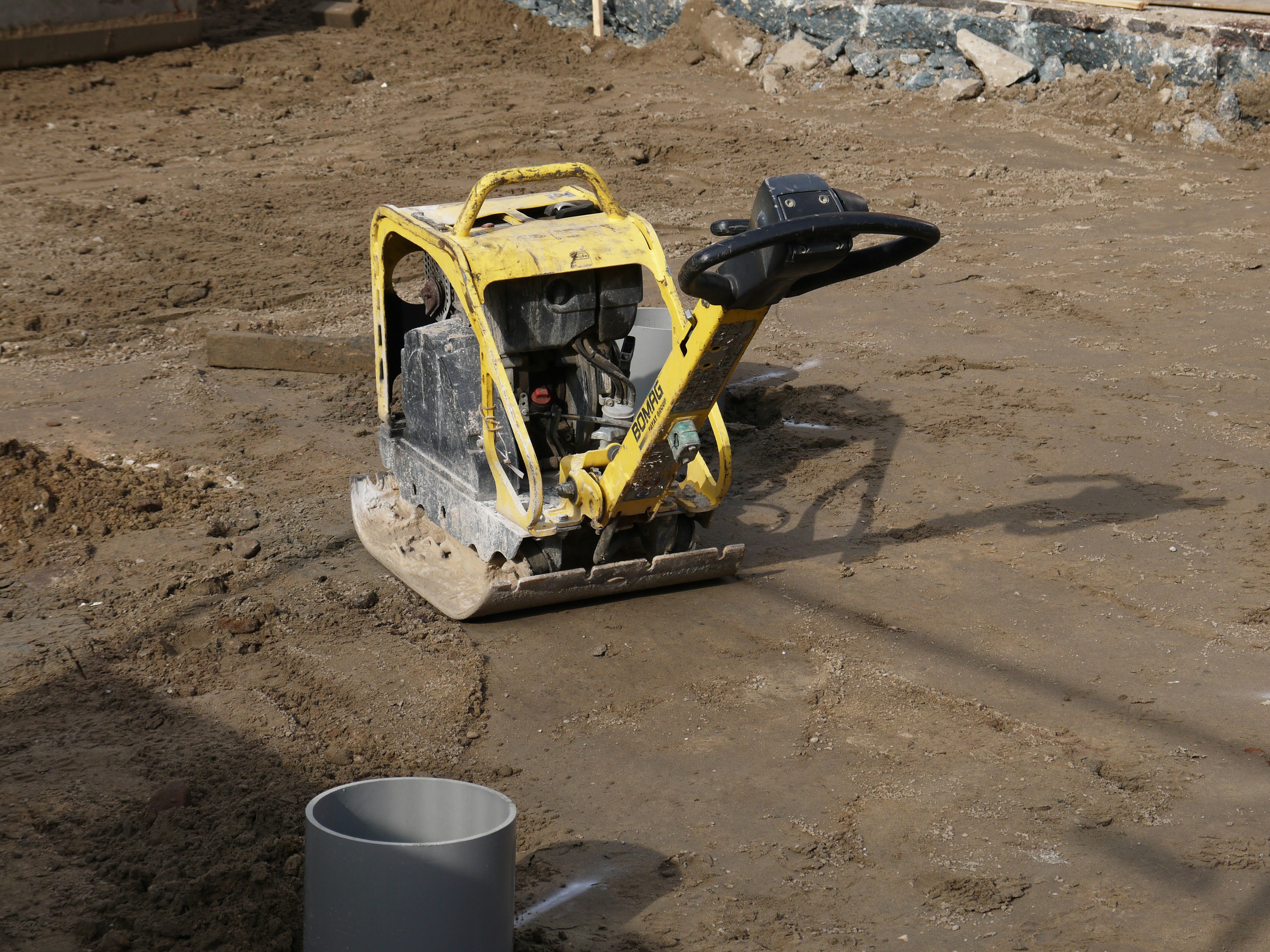 Compact yellow plate compactor resting on a construction site with a gray pipe nearby on the dirt surface.