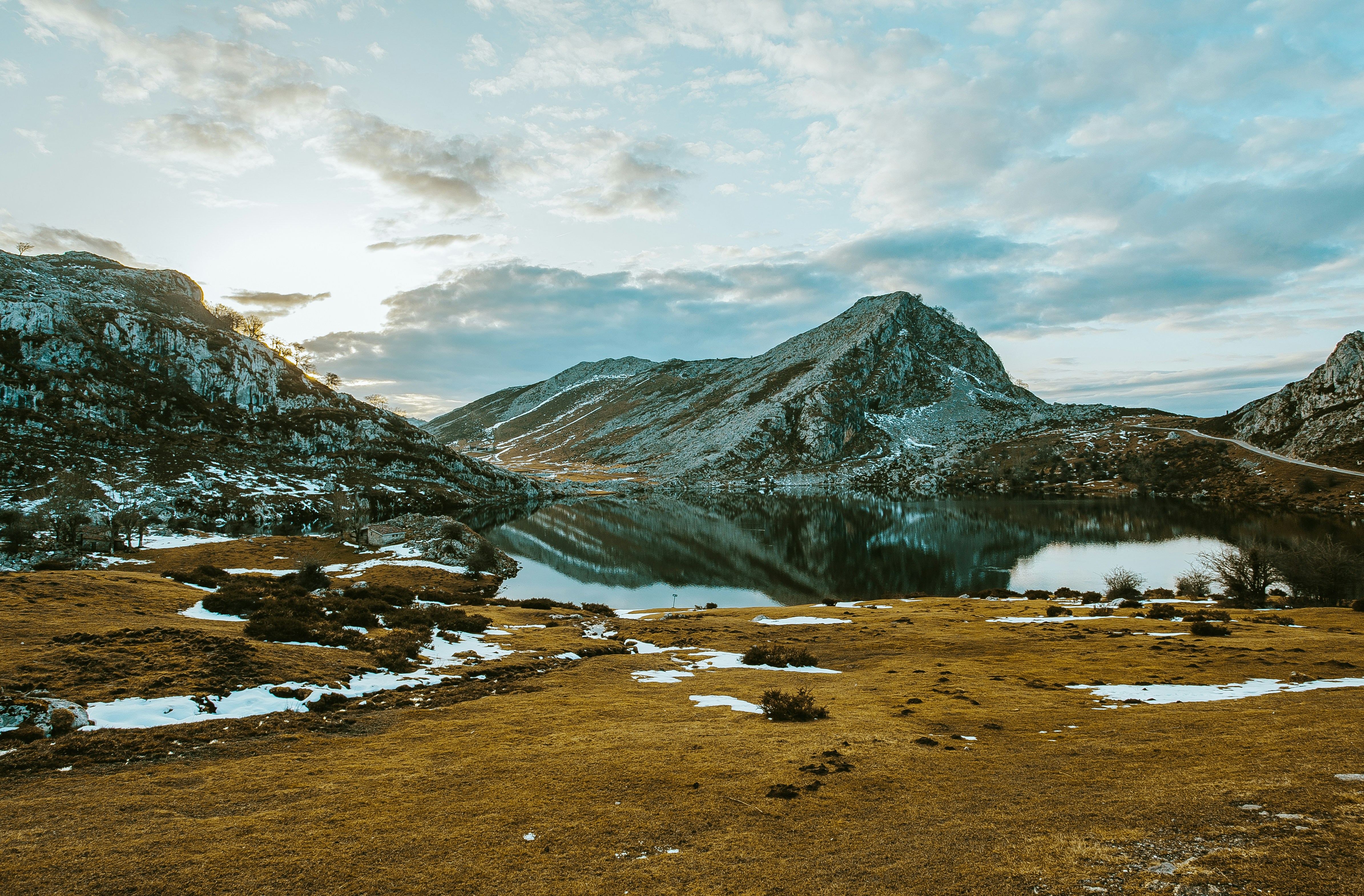 Snow-dusted mountains reflecting in a tranquil lake, framed by golden grasslands under a cloudy sky.