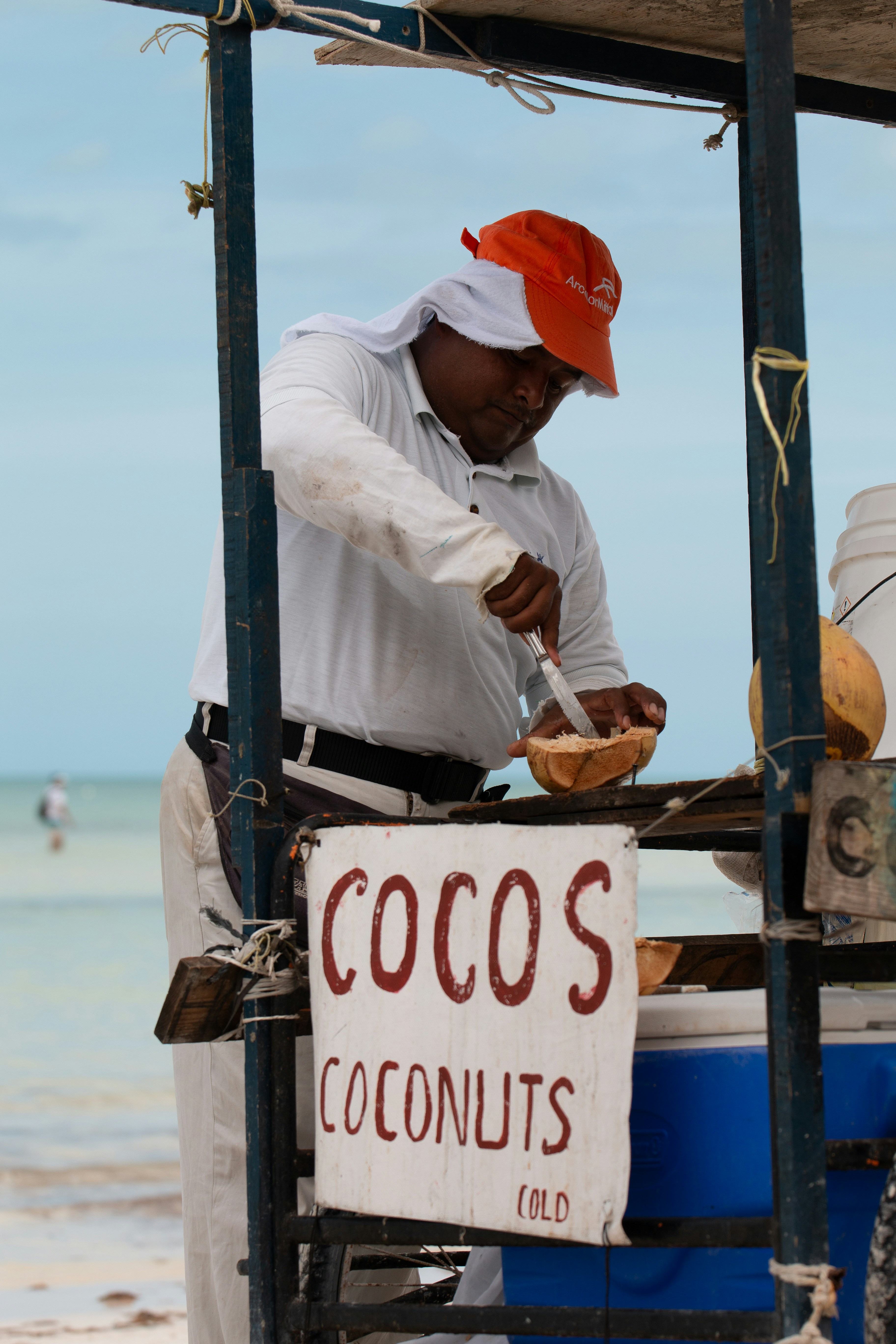 Coconut Vendor at the Seashore: Hands at Work Beneath a Bright Orange Hat