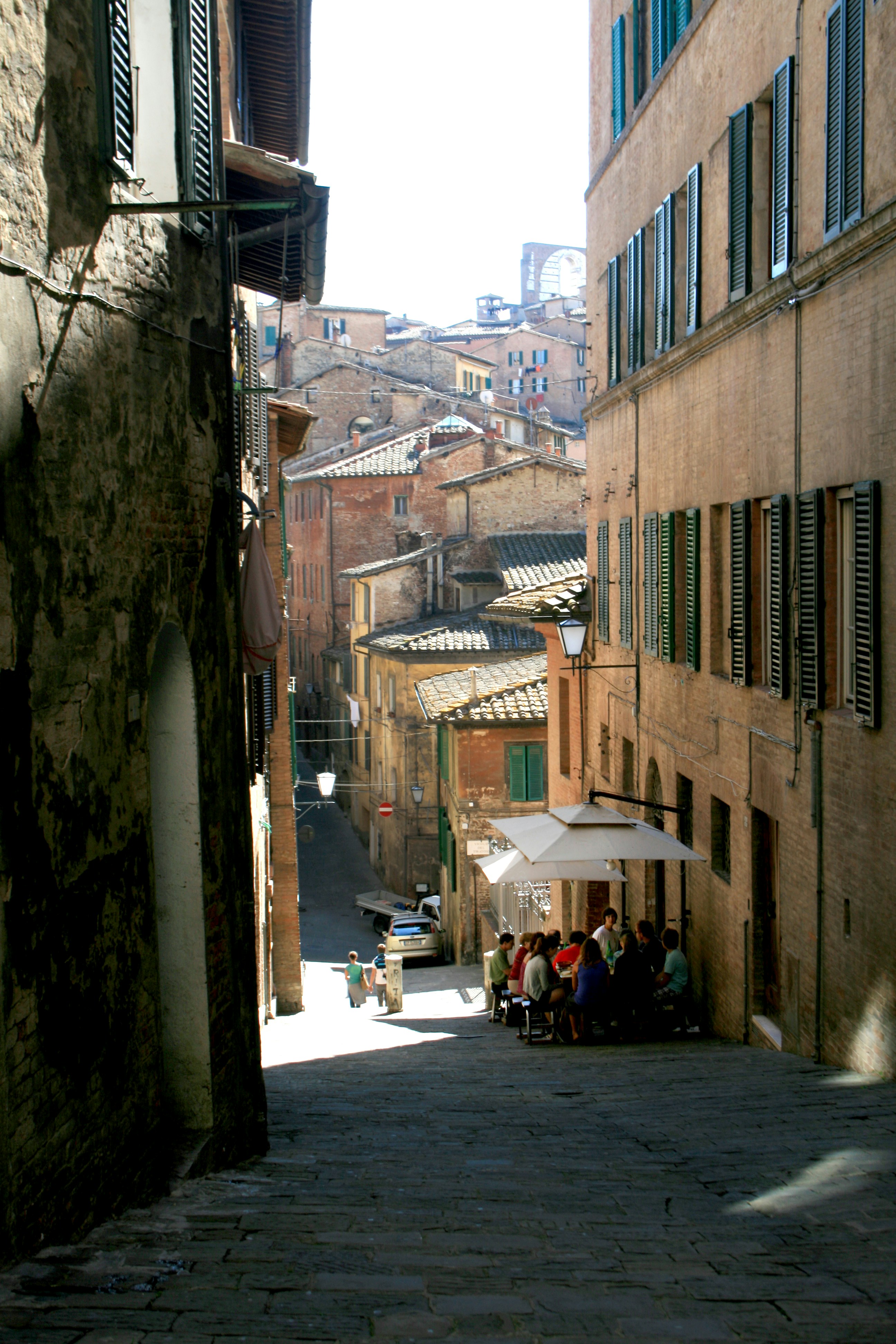 Charming narrow street in Siena leading to a lively outdoor café, surrounded by historic buildings and sunlit rooftops.