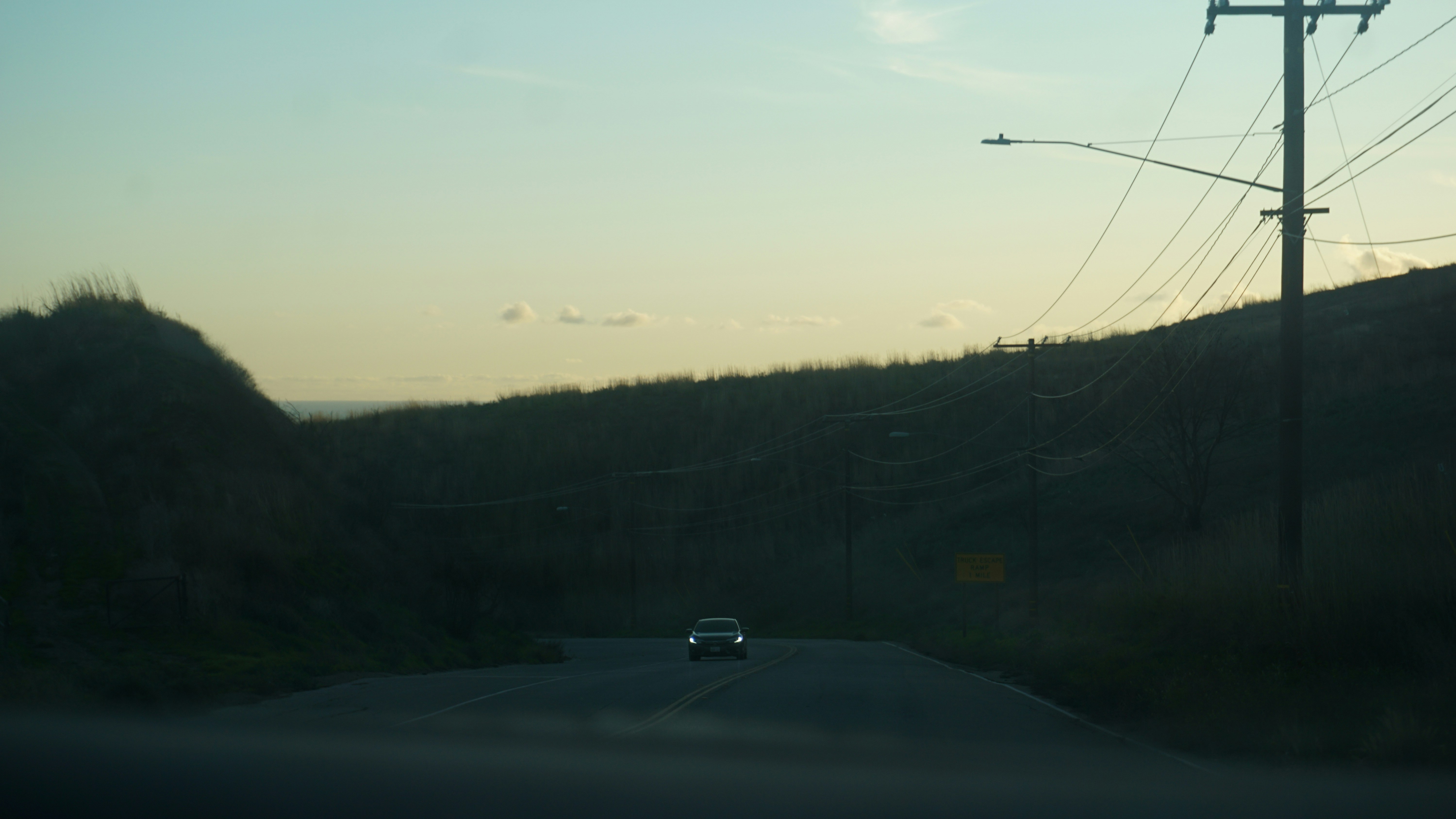 A solitary car navigates a winding road surrounded by rolling hills at dusk, with power lines framing the scene. The fading light casts a serene ambiance.