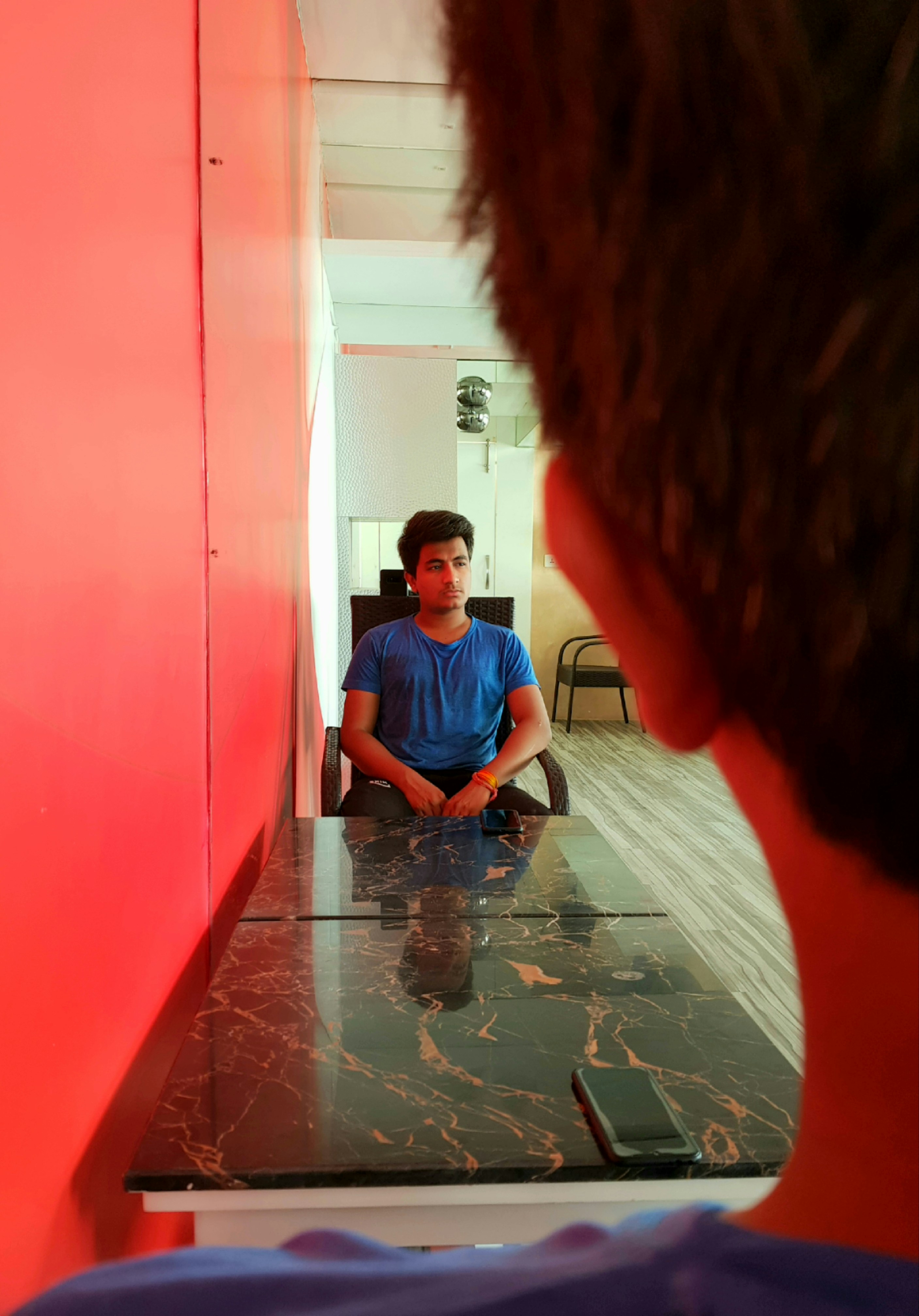 A young man in a blue shirt sits at a table, gazing ahead, while another figure partially frames the scene from the foreground. The vibrant red wall adds a striking backdrop.
