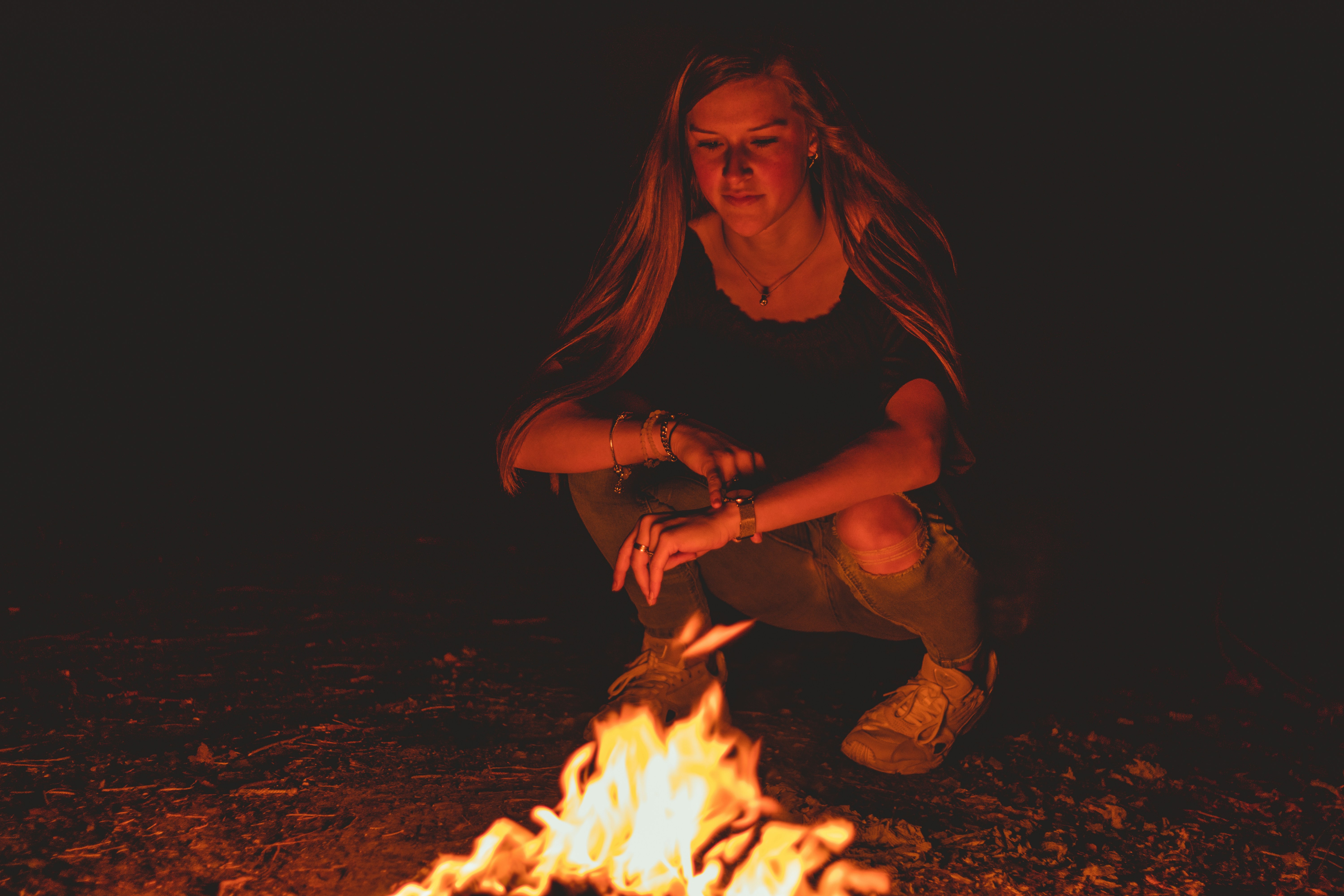 Woman crouches beside a flickering campfire, adjusting bracelets as warm flames illuminate her face. Night air surrounds the scene, emphasizing the intimate, contemplative moment.