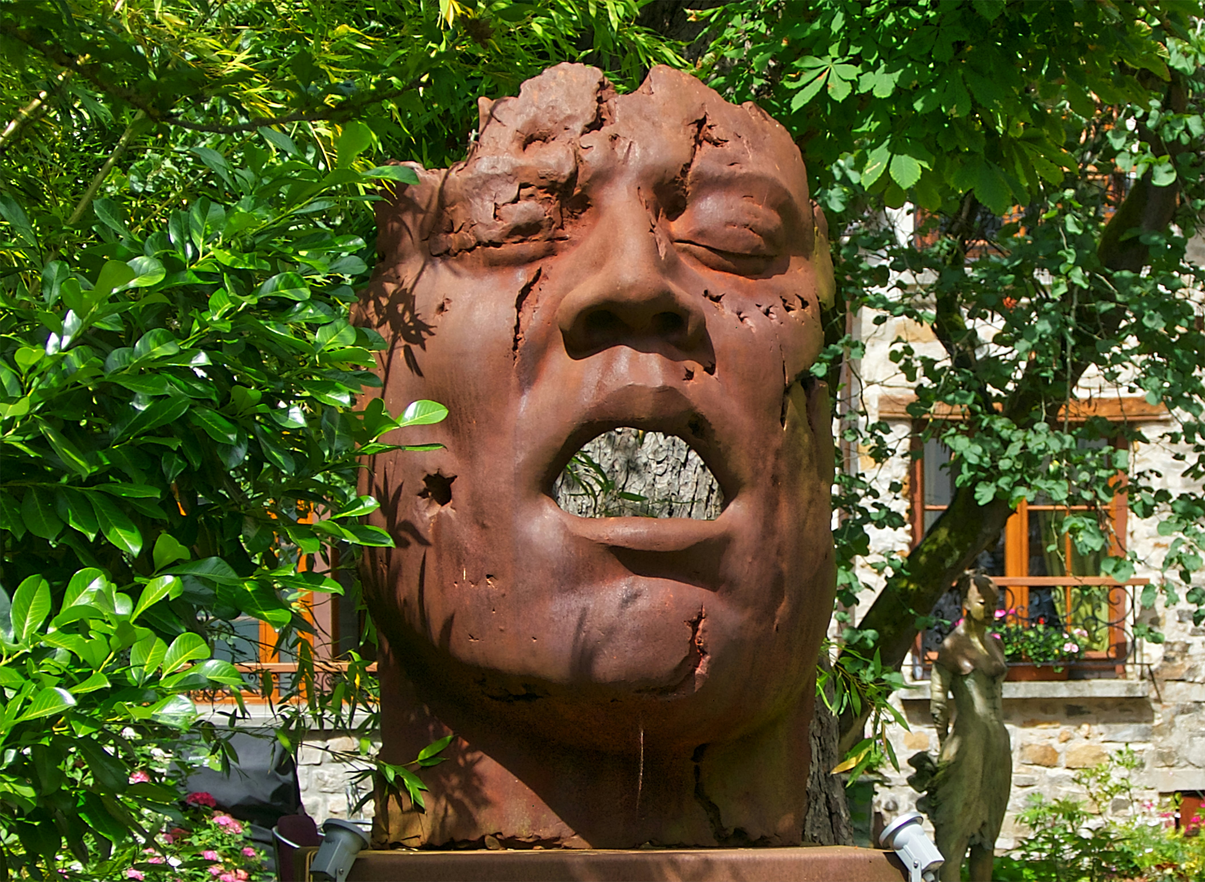 A large, rusted sculpture of a face expressing anguish, surrounded by lush greenery. The piece stands as a striking contrast to the natural setting.