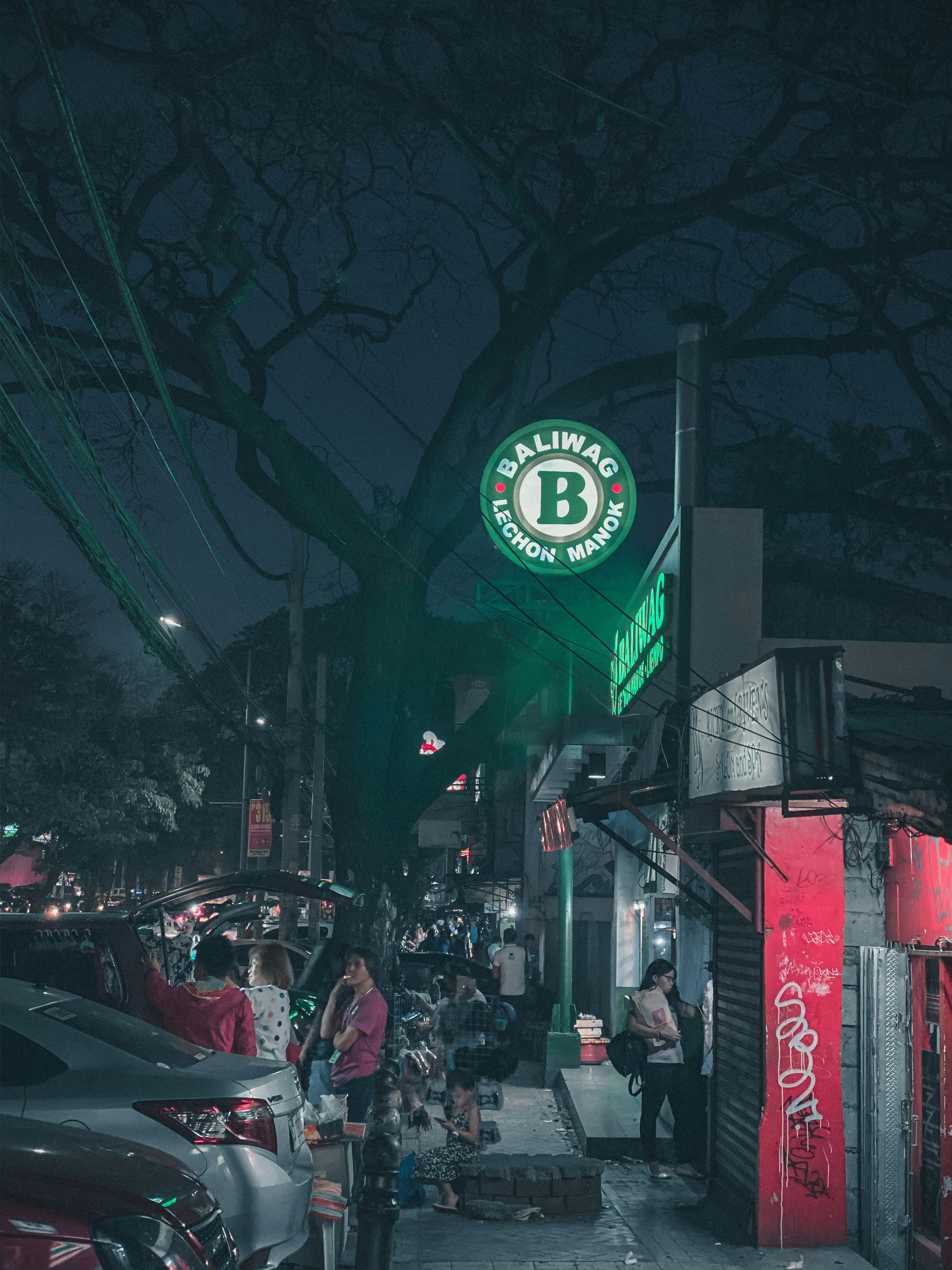 A vibrant street scene featuring the illuminated sign of Baliwag Lechon Manok, with people mingling in the background and a glimpse of parked cars in the foreground.
