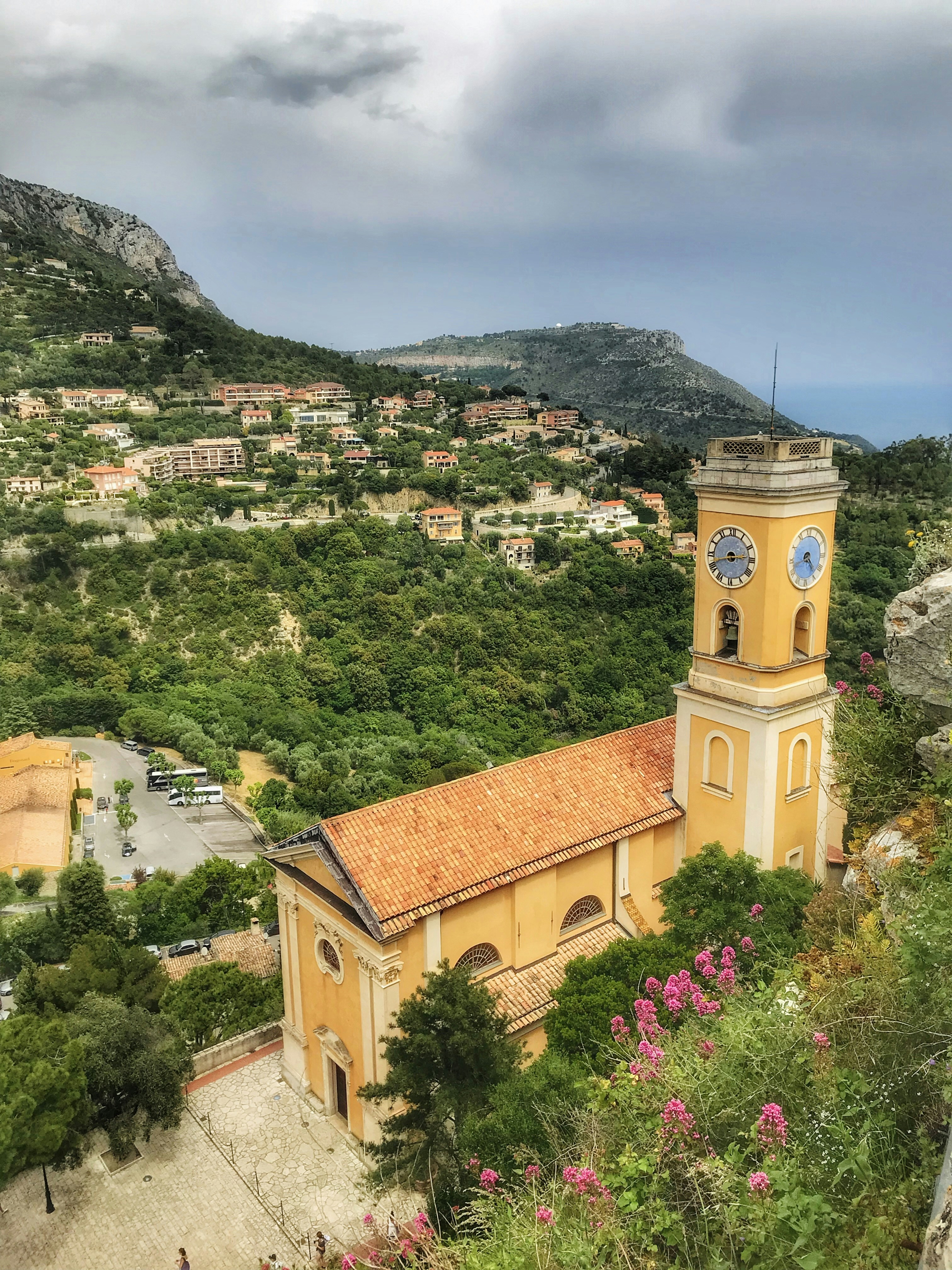Historic yellow church tower amidst lush greenery and distant sea view in Eze Village.