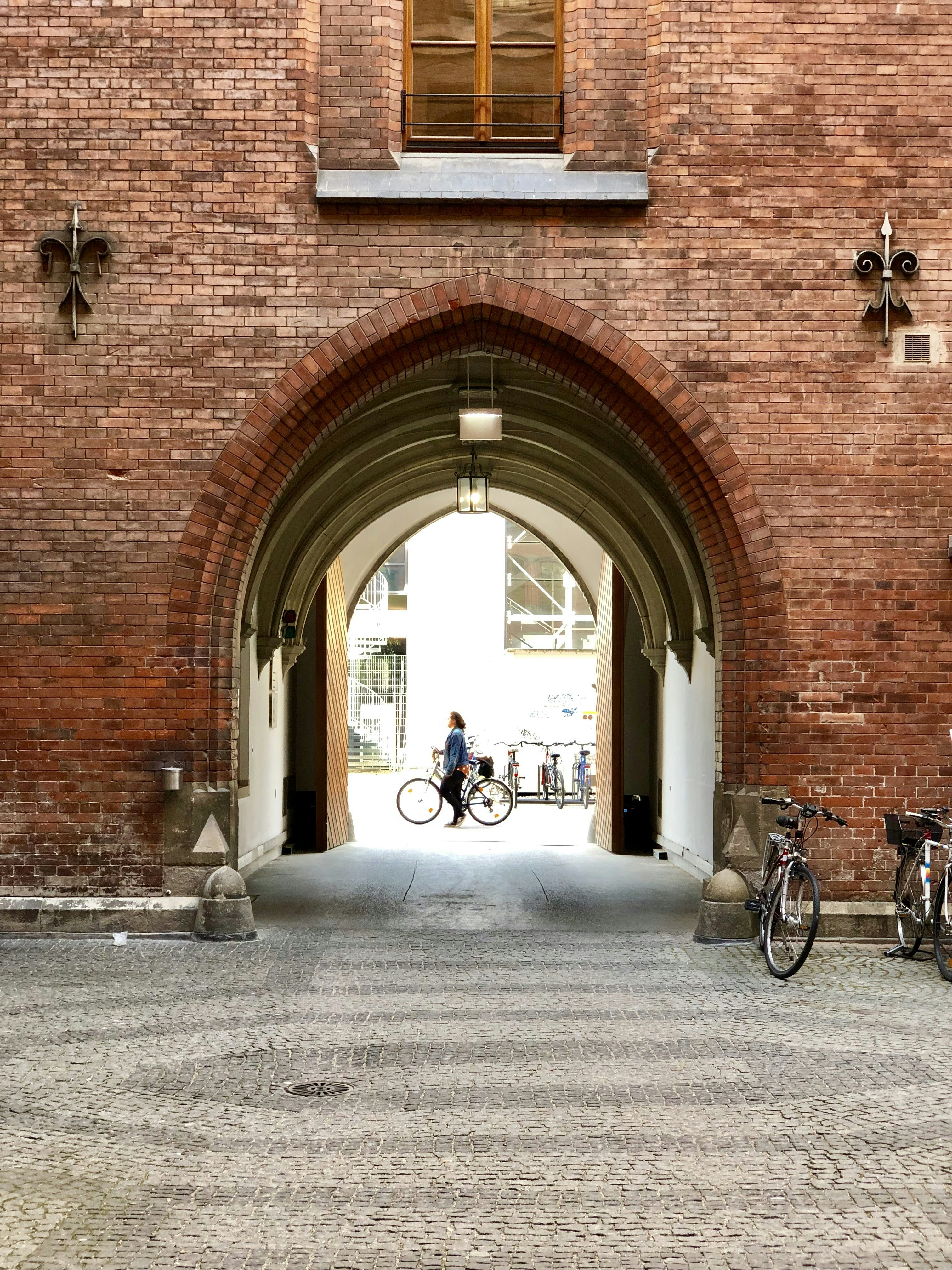 Person walking a bicycle through a brick archway into a sunlit courtyard.