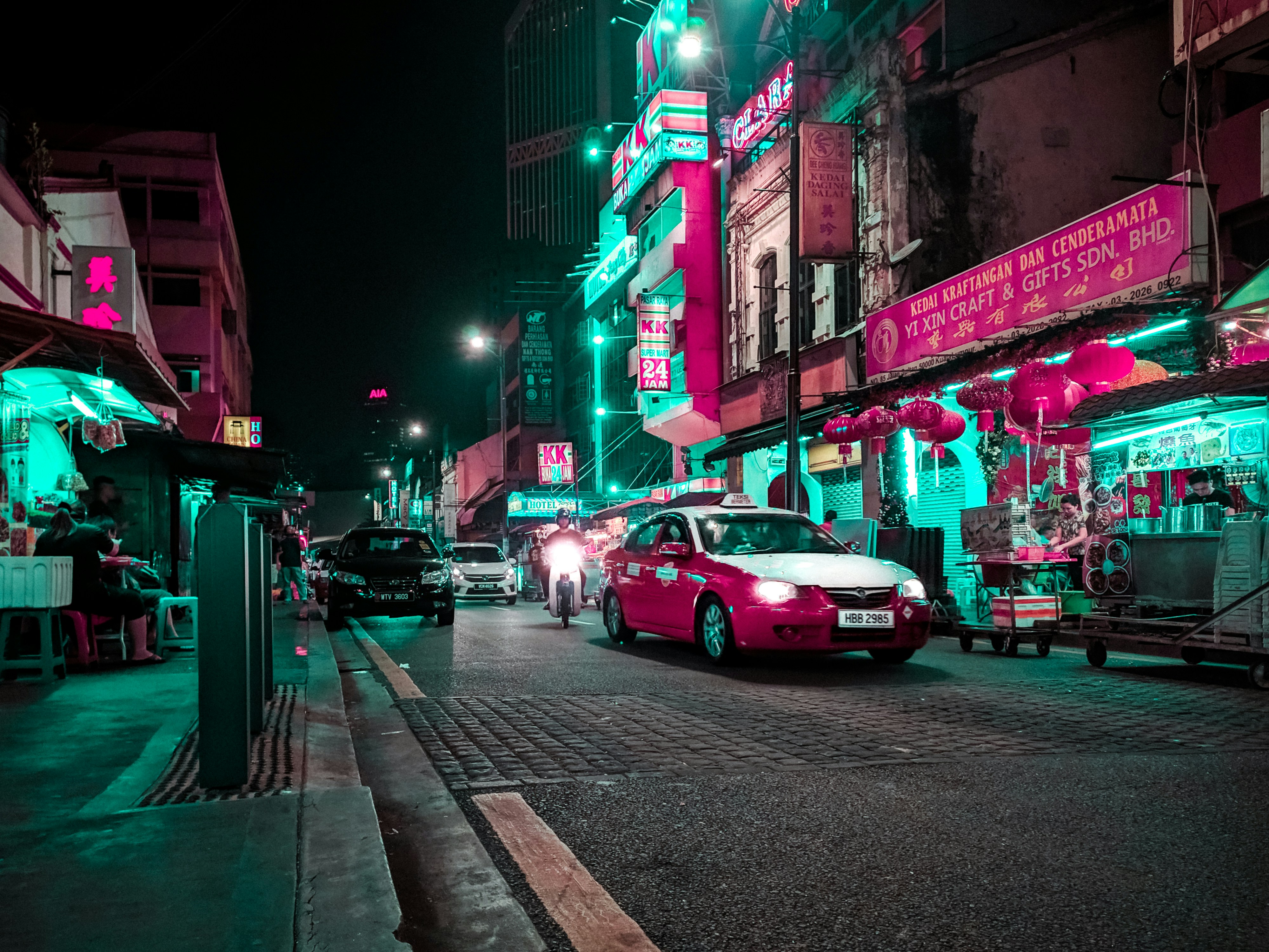 Vibrant neon lights illuminate a bustling night market street with a car in motion.