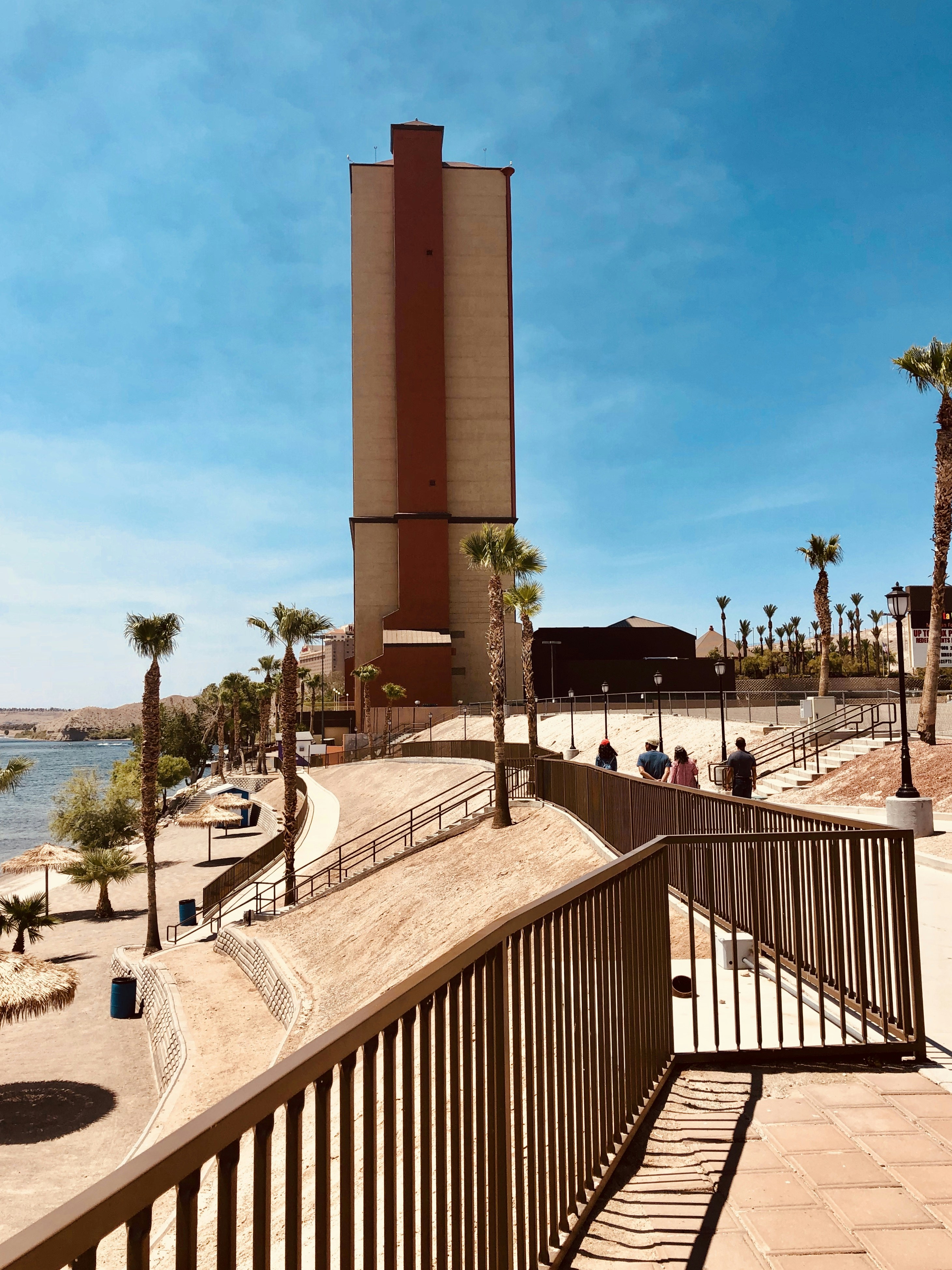 Riverside walkway lined with palm trees and a tall building in the background, with people enjoying a leisurely stroll. The scene captures a blend of nature and urban architecture.
