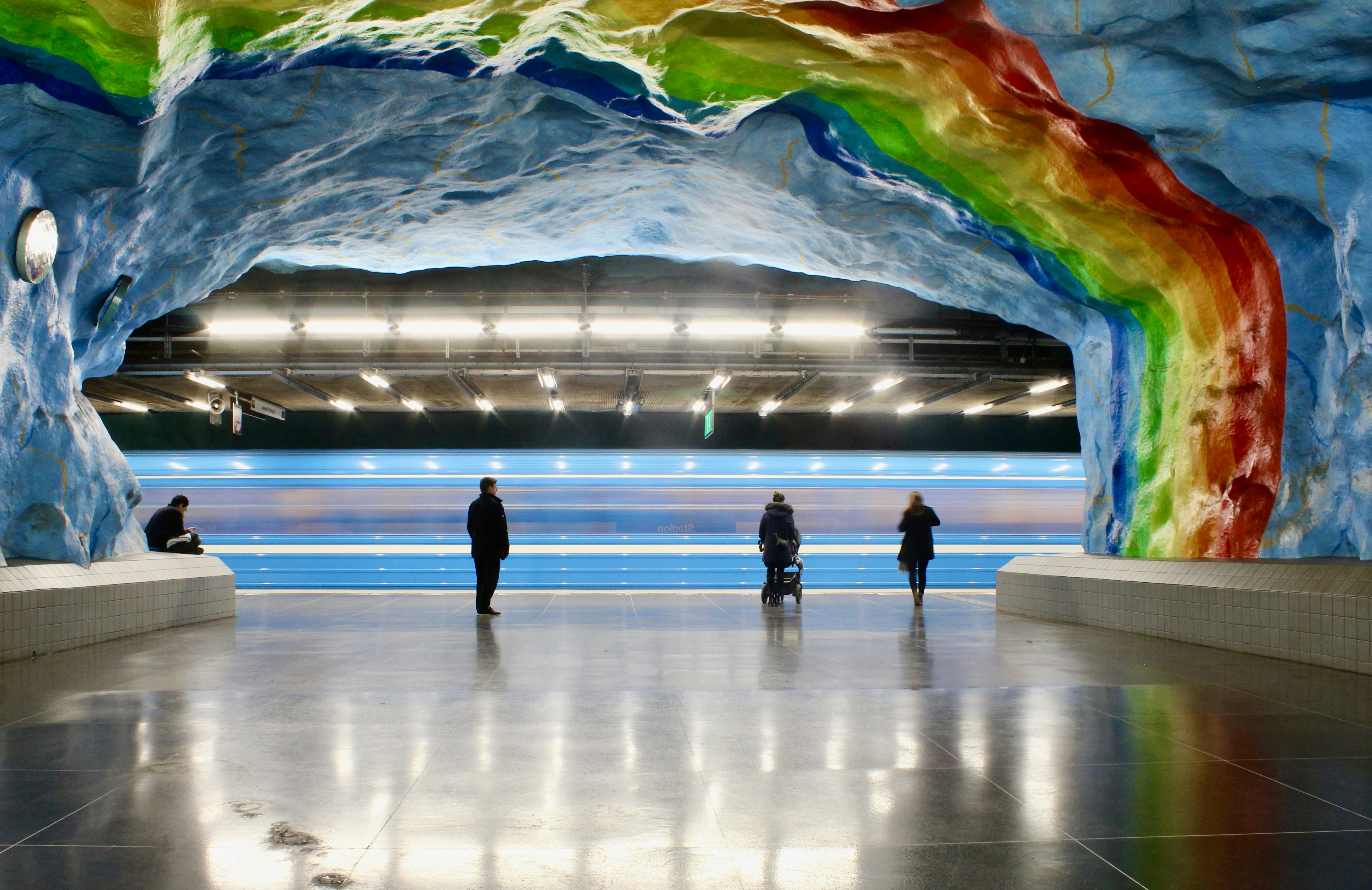 Vibrant rainbow-colored rock formations in a Stockholm metro station with blurred train motion and silhouetted figures.