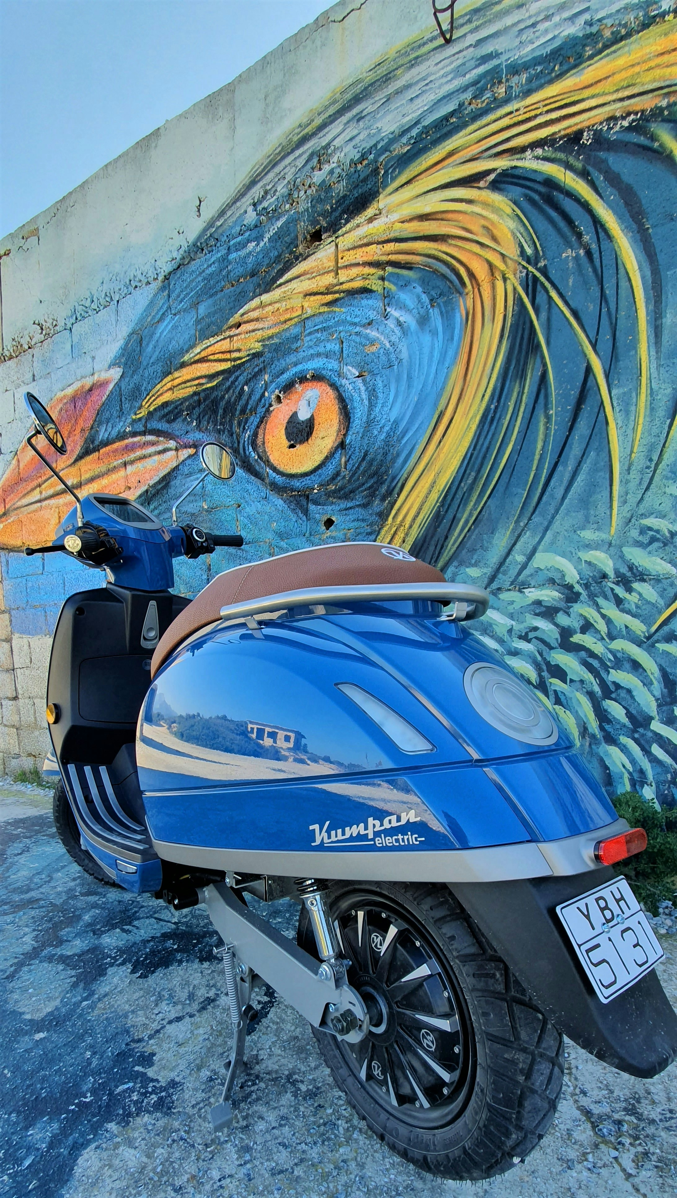 A vibrant blue electric scooter parked beside a colorful mural featuring a bird's eye. The juxtaposition highlights urban art and modern transportation.