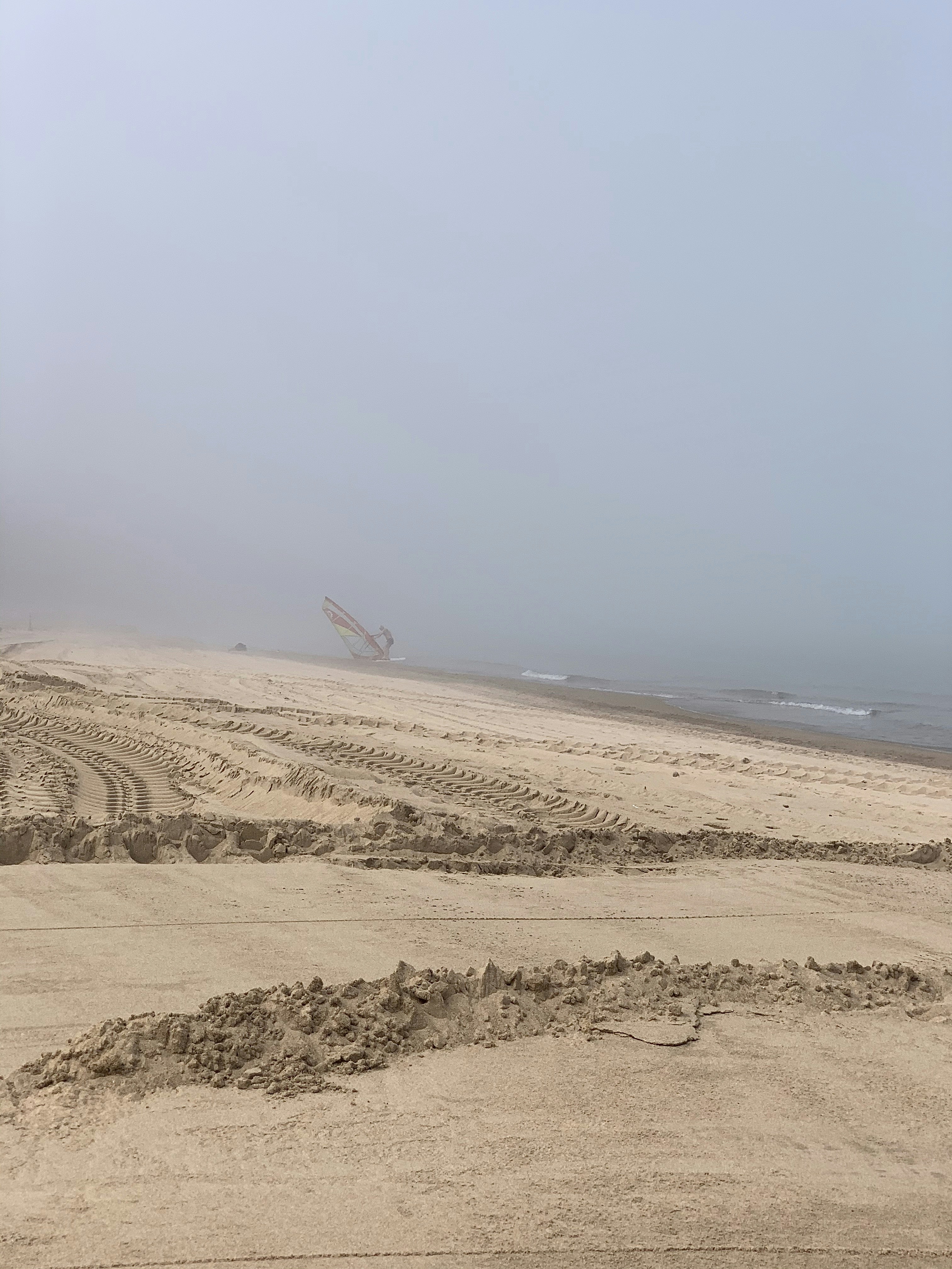 A serene beach scene enveloped in fog, with gentle waves lapping at the shore and distant silhouettes of fishing boats. The sand shows traces of tire tracks leading into the mist.