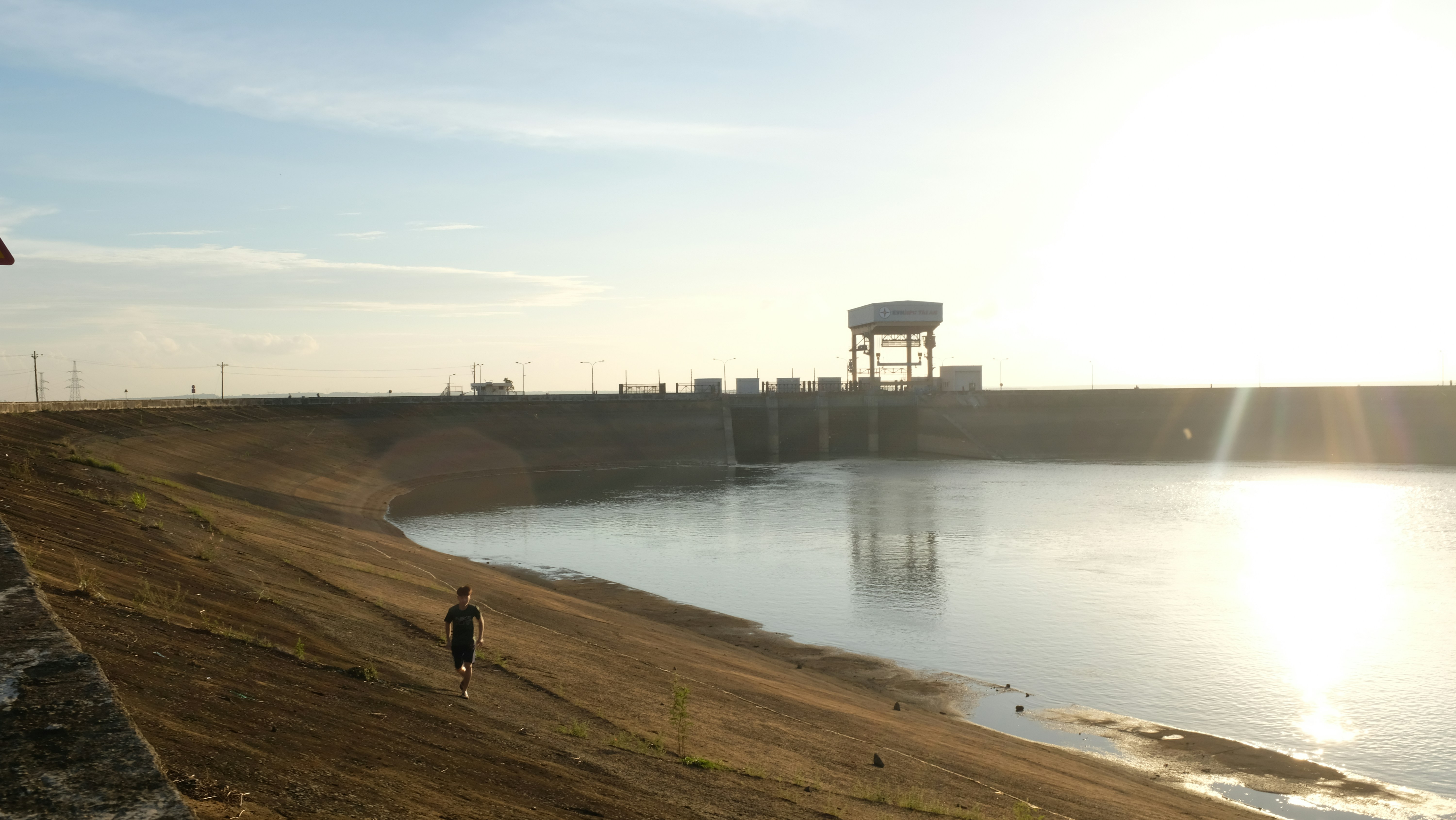 A solitary figure strolls along a dry embankment beside a calm reservoir, with a water tower and dam structure in the background. Sunlight glints off the water's surface.