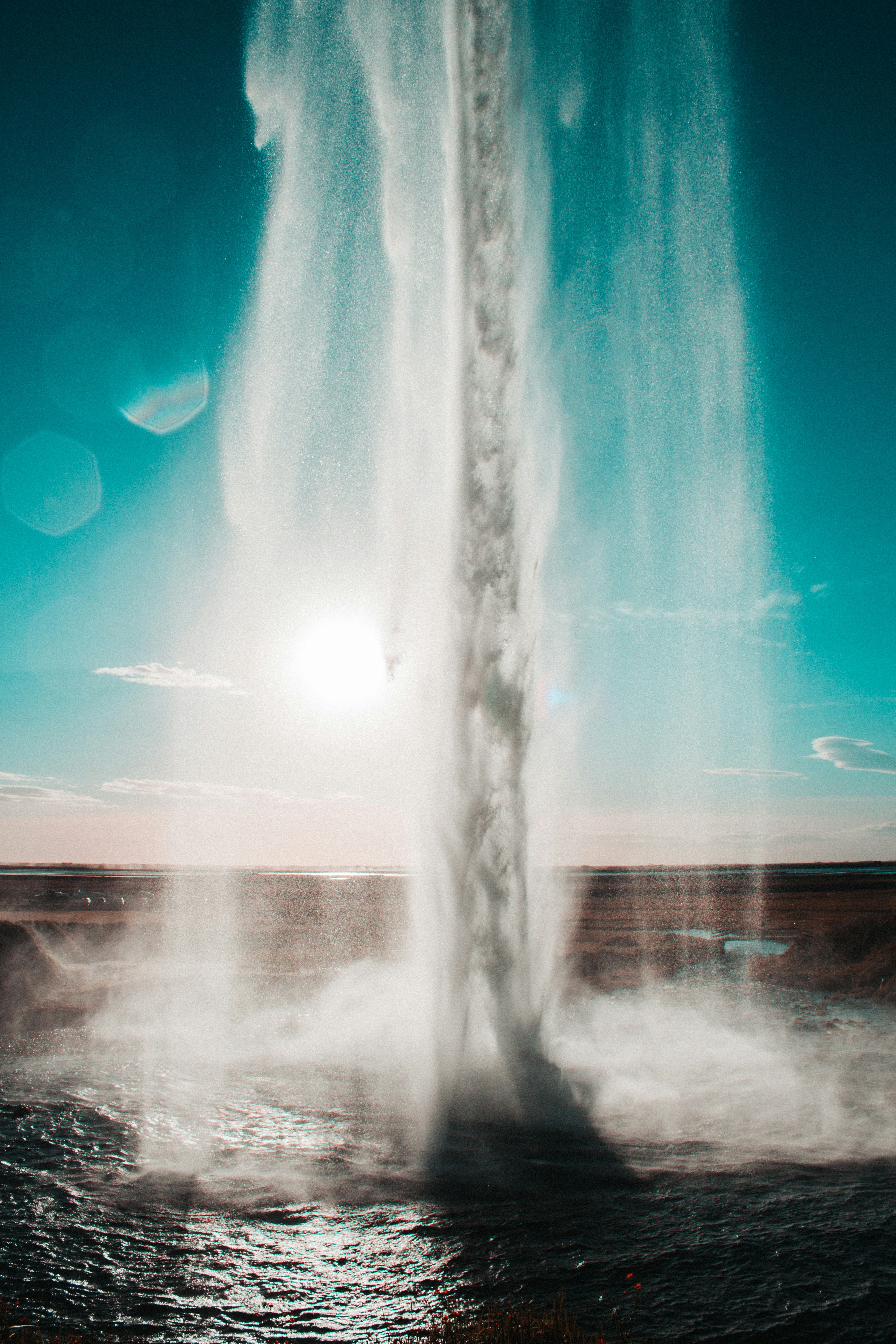 Geyser erupting with a powerful spray against a backdrop of a bright sun and clear blue sky.