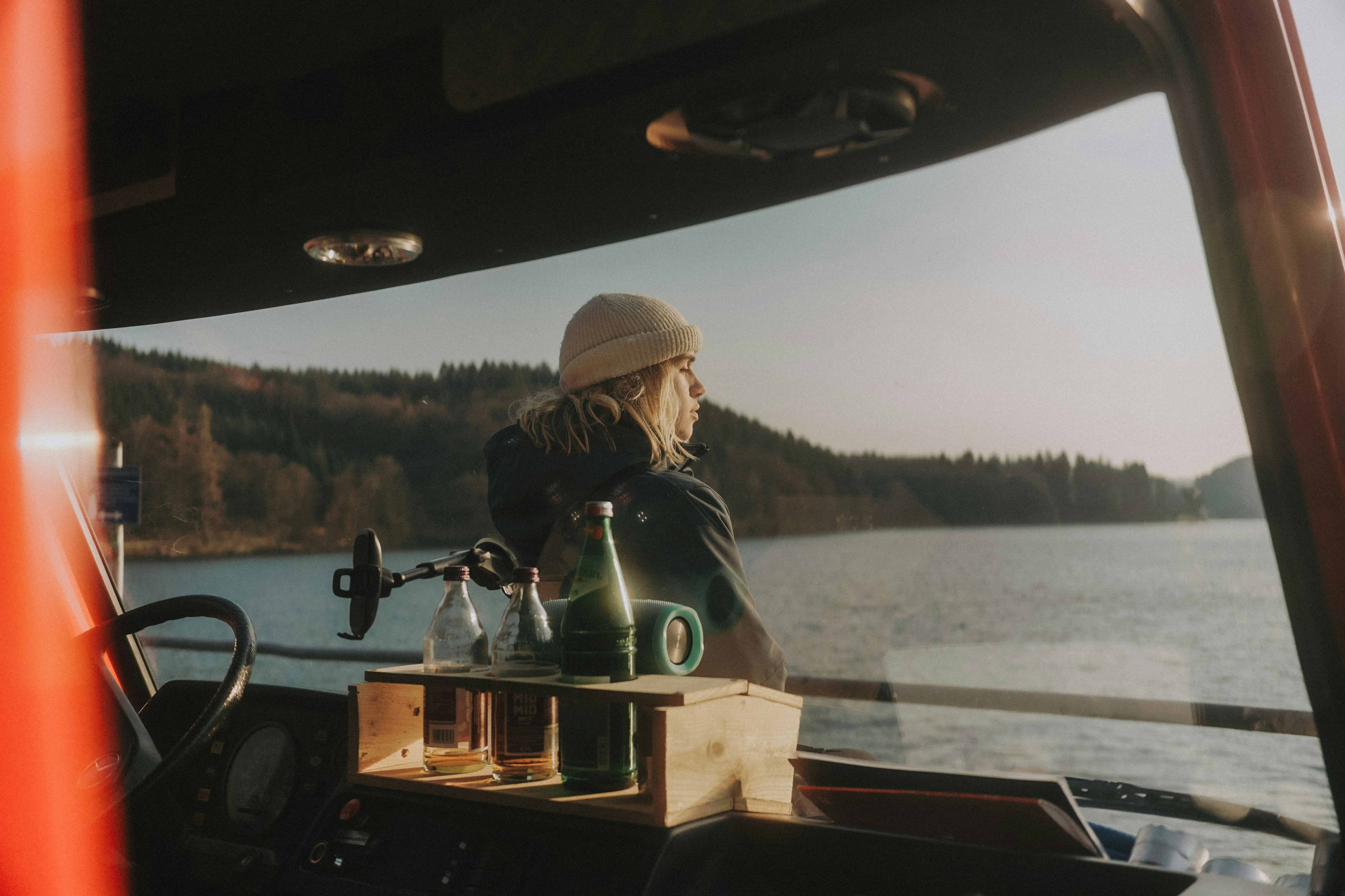 A person gazes out over a tranquil lake from inside a boat, framed by the cabin’s interior. Sunlight casts a warm glow on the scene.