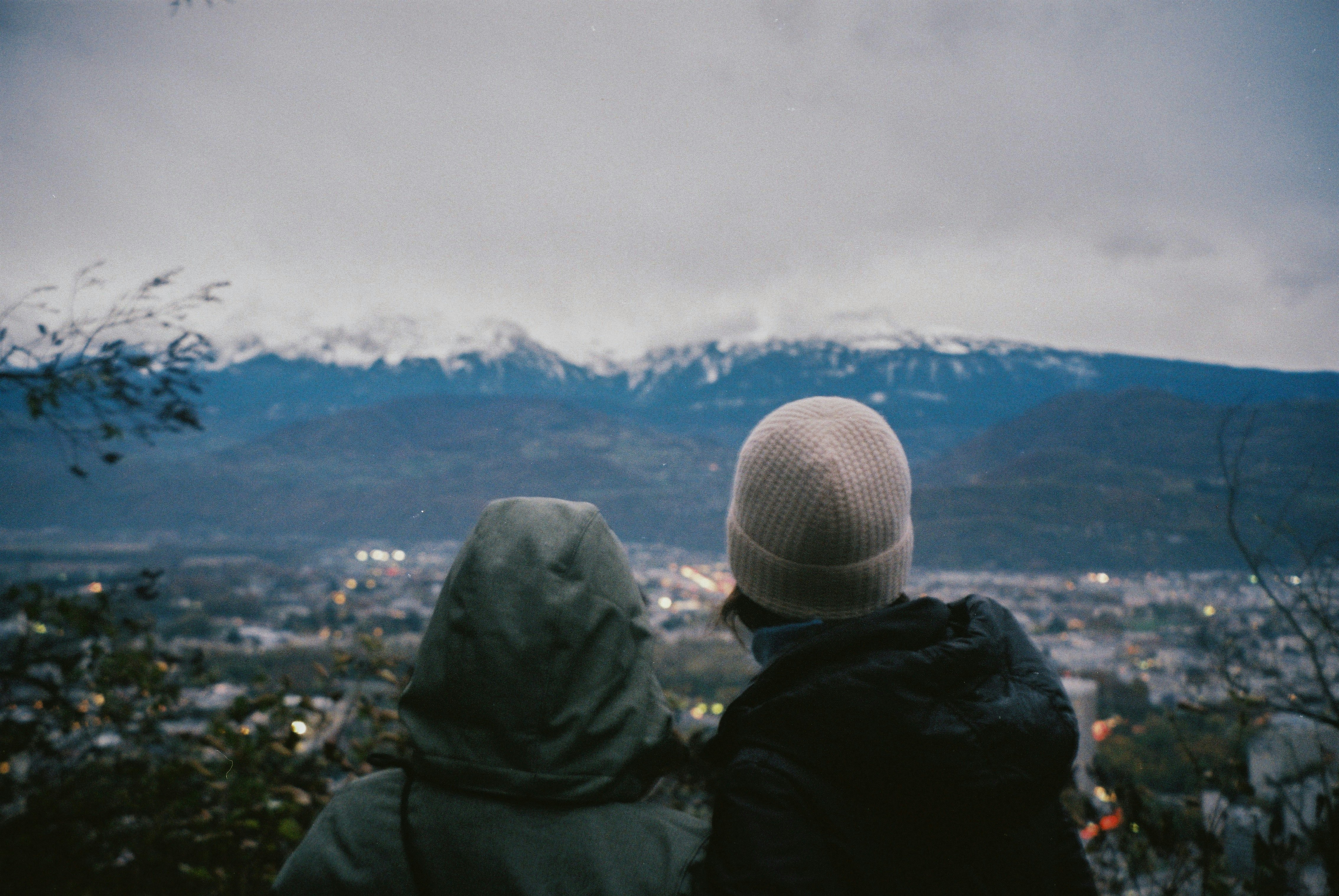 Two people in winter clothing overlook a sweeping mountain landscape under a cloudy sky.