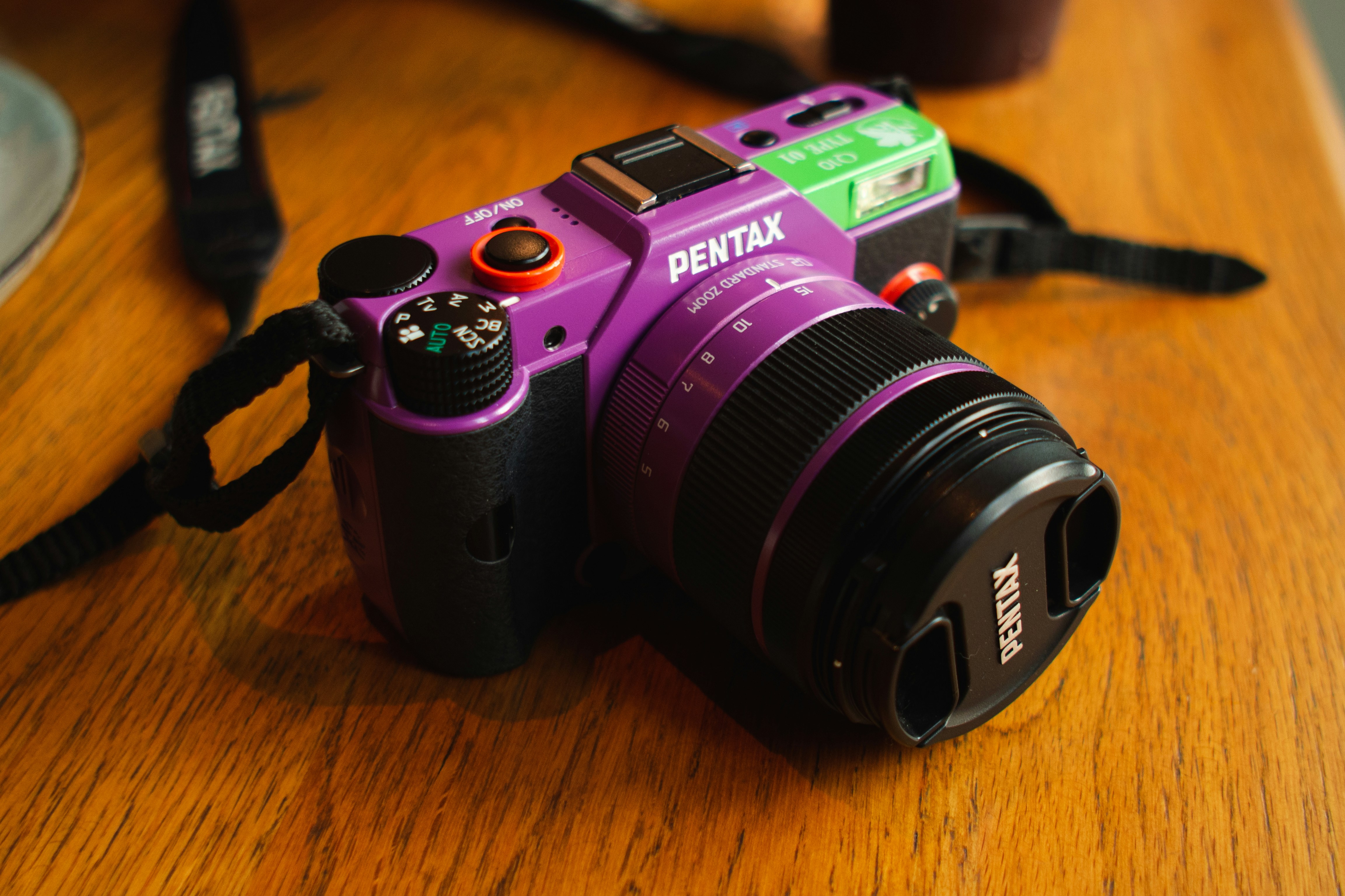 Colorful Pentax camera resting on a wooden surface, showcasing its unique design and lens. The scene captures the essence of photography gear in a casual setting.