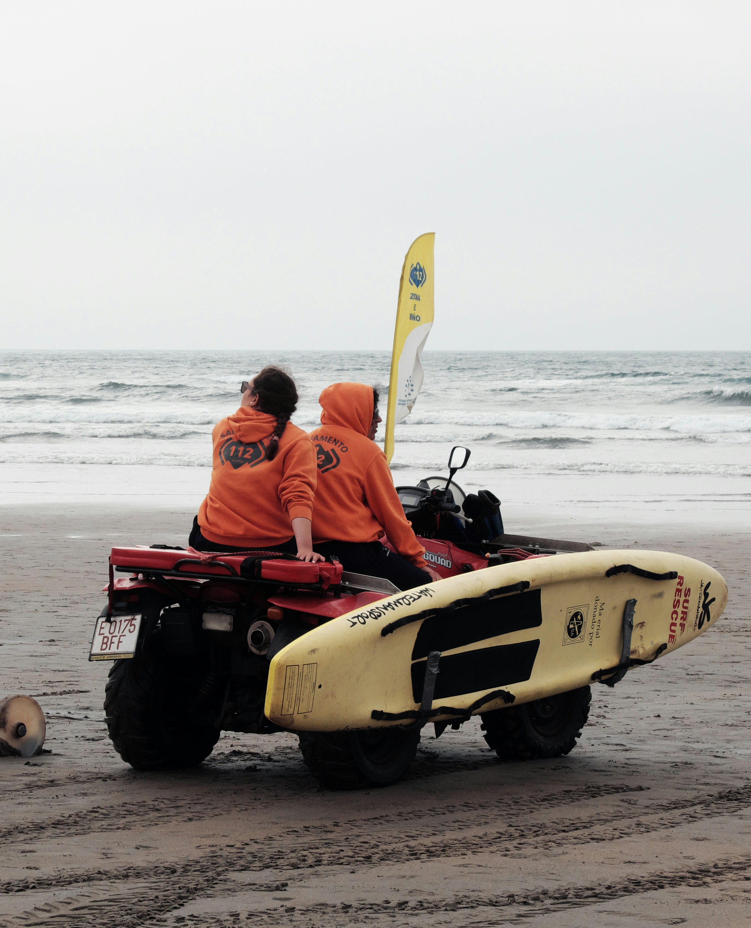 Two Orange-Hooded Riders Secure a Yellow Surfboard Beside a Beachfront ATV