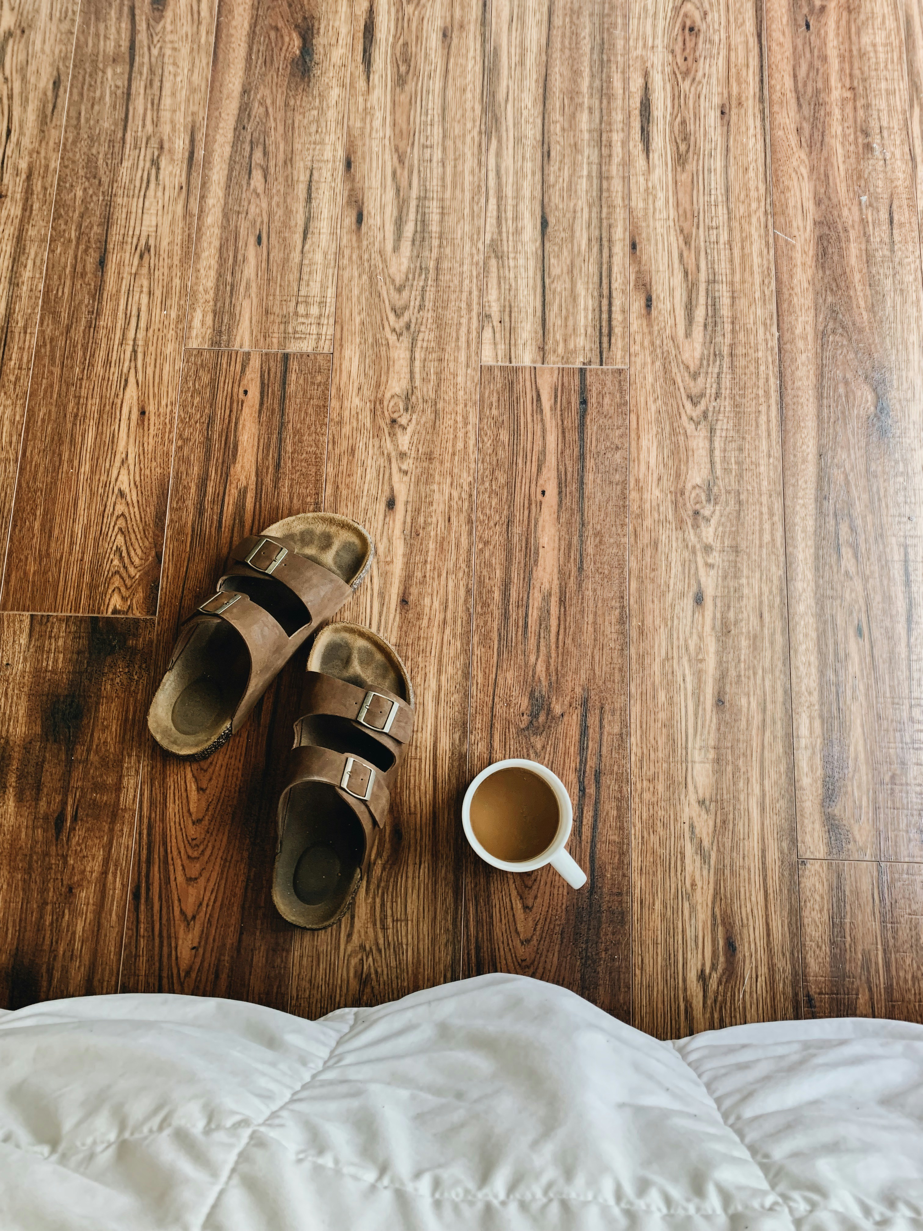 This image captures the essence of a tranquil morning with a pair of well-worn sandals and a steaming cup of coffee resting on a rich, textured wooden floor. The warm tones of the wood complement the earthy brown of the sandals, while the inviting white duvet adds a touch of comfort to the composition. The natural lighting softly highlights the scene, creating a calm and inviting atmosphere that emphasizes the simplicity and coziness of a homebody's morning ritual.