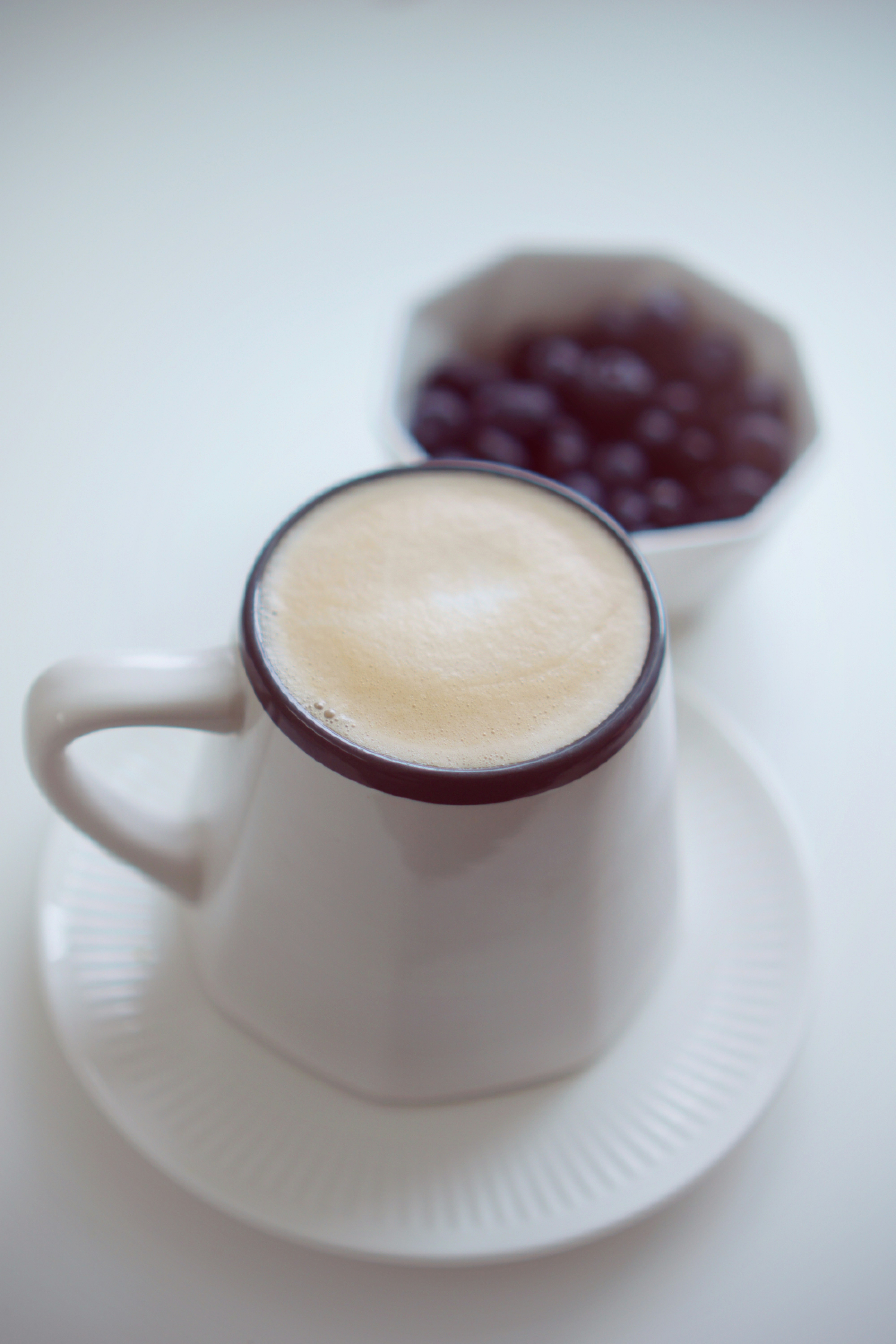 A white coffee mug filled with frothy beverage sits on a plate beside a bowl of dark berries, set against a light background.