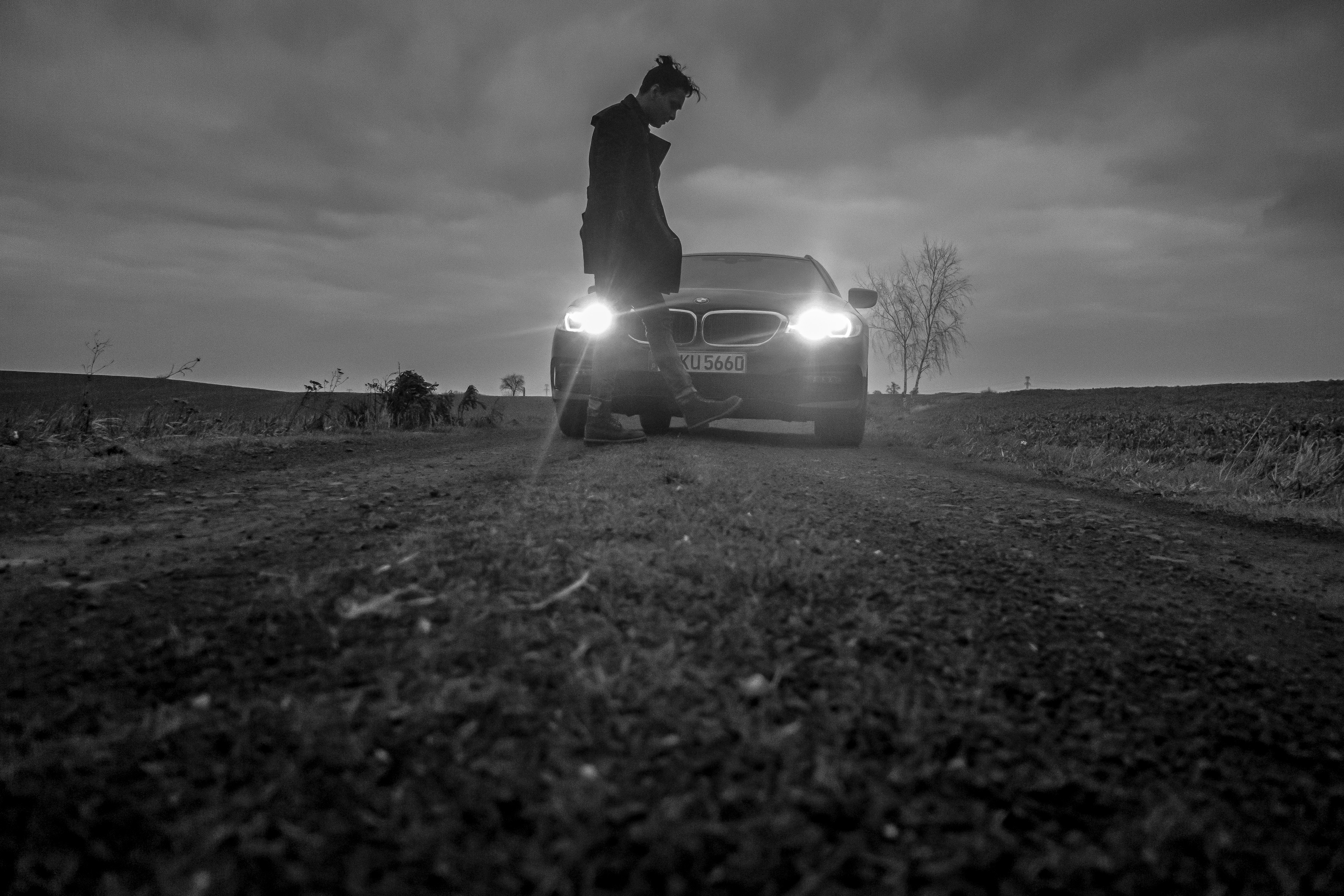 A figure in a dark coat stands beside a sleek car on a desolate dirt road, under an overcast sky. The scene evokes a sense of solitude and introspection.