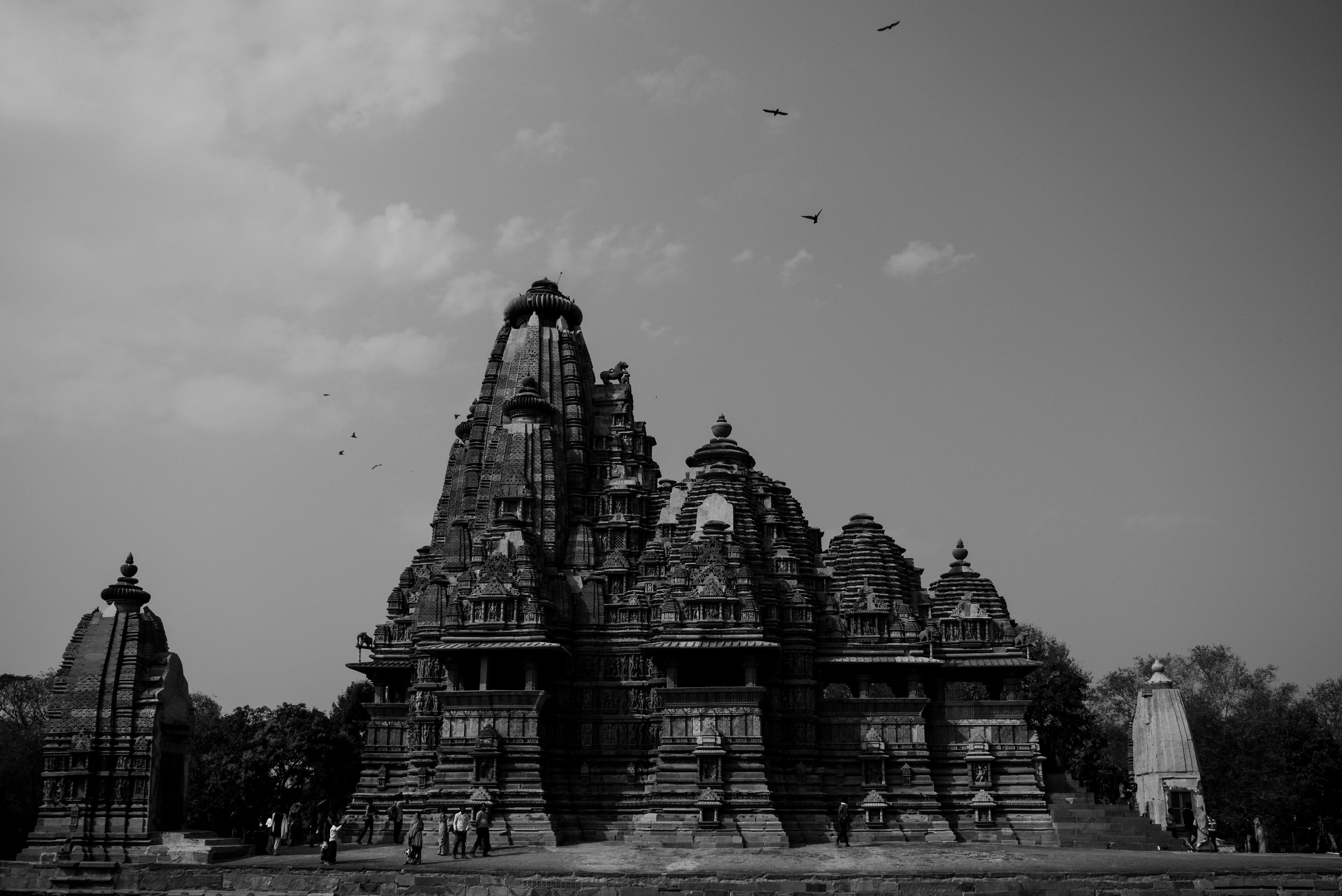 Ancient temple structures stand under a partly cloudy sky, with birds soaring above.