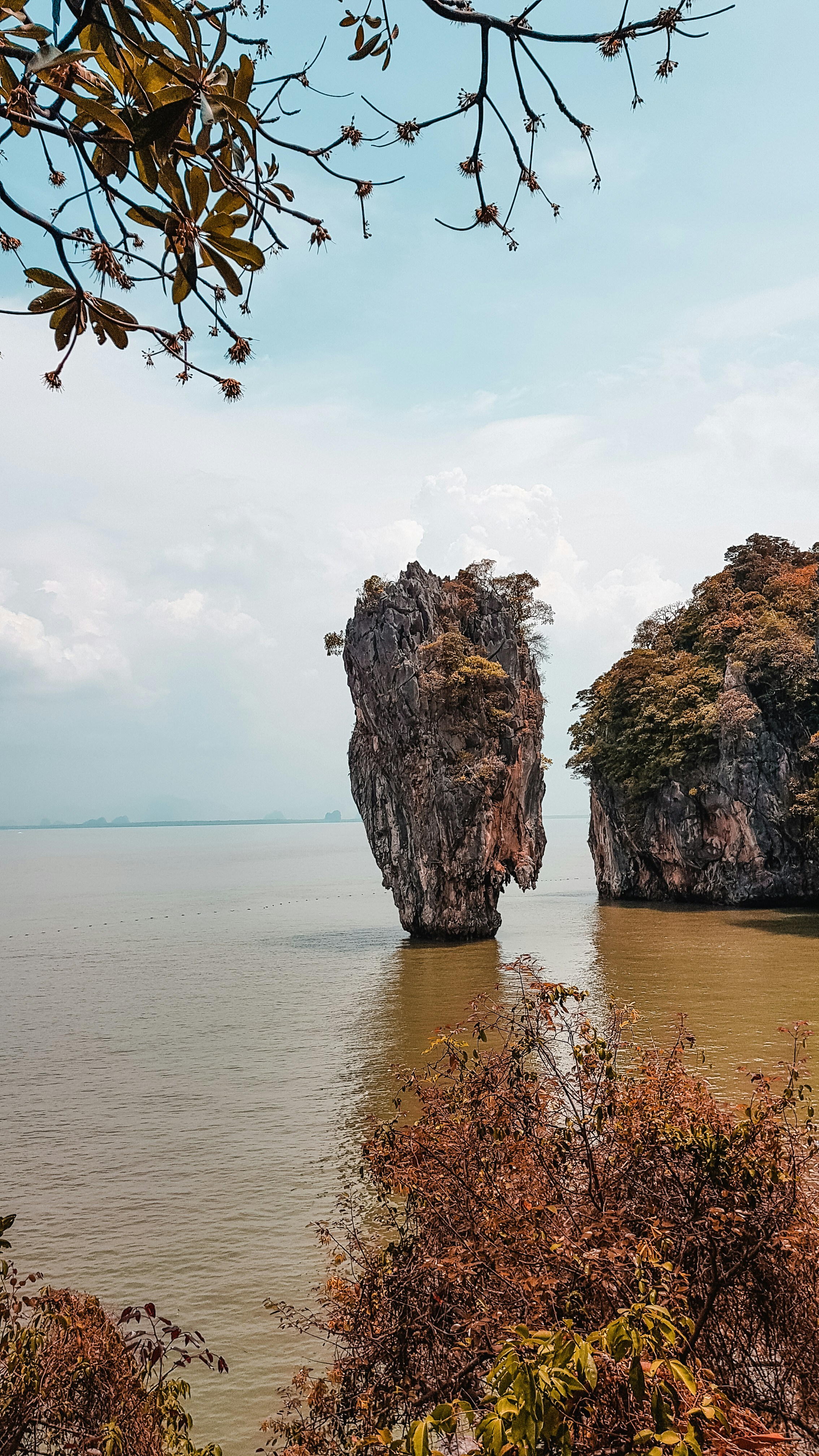 A towering rock formation emerges from calm waters, flanked by lush greenery and a serene sky. The scene captures the tranquil beauty of nature's artistry.
