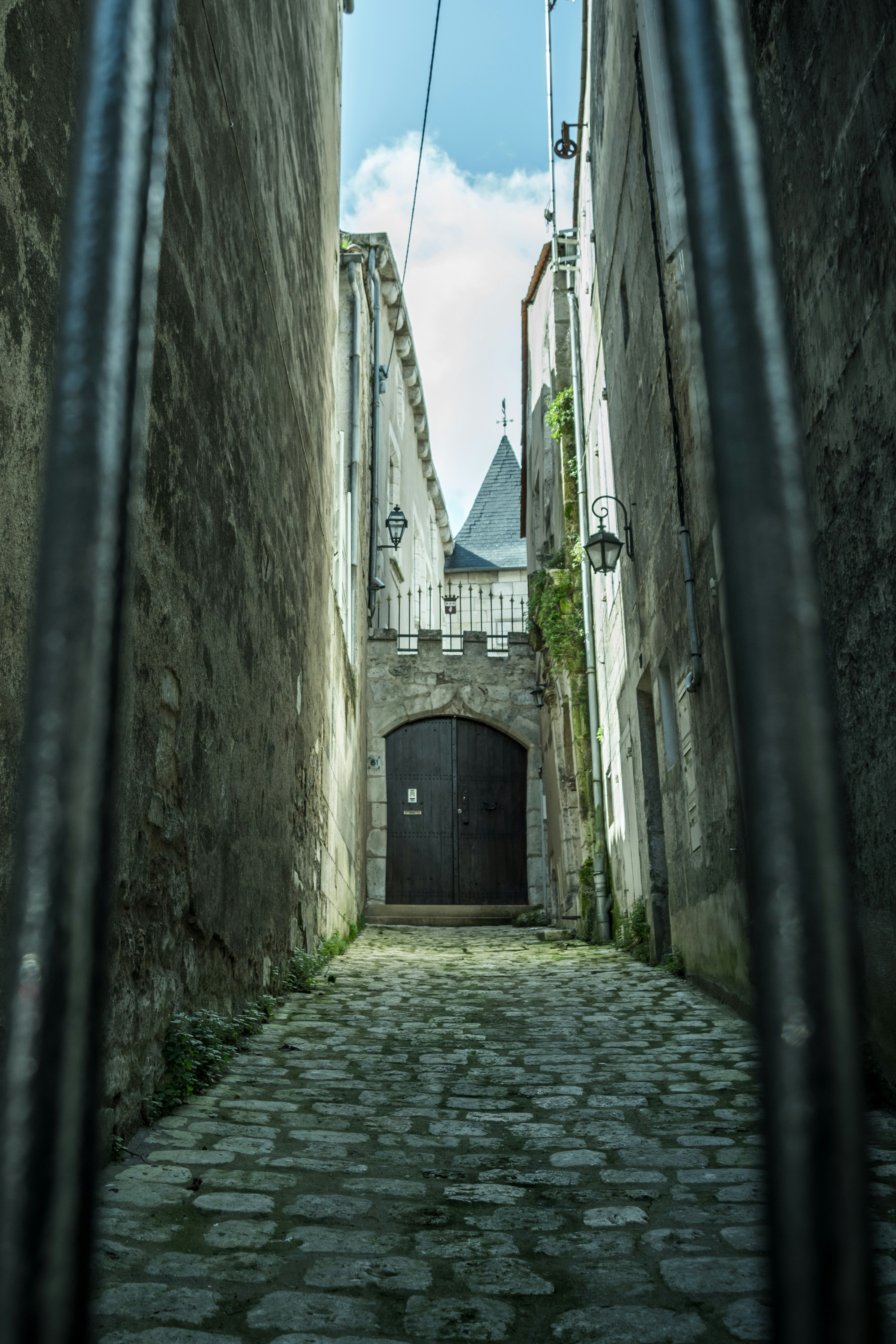 Narrow cobblestone alleyway flanked by tall weathered walls leading to an arched wooden door under a bright sky.