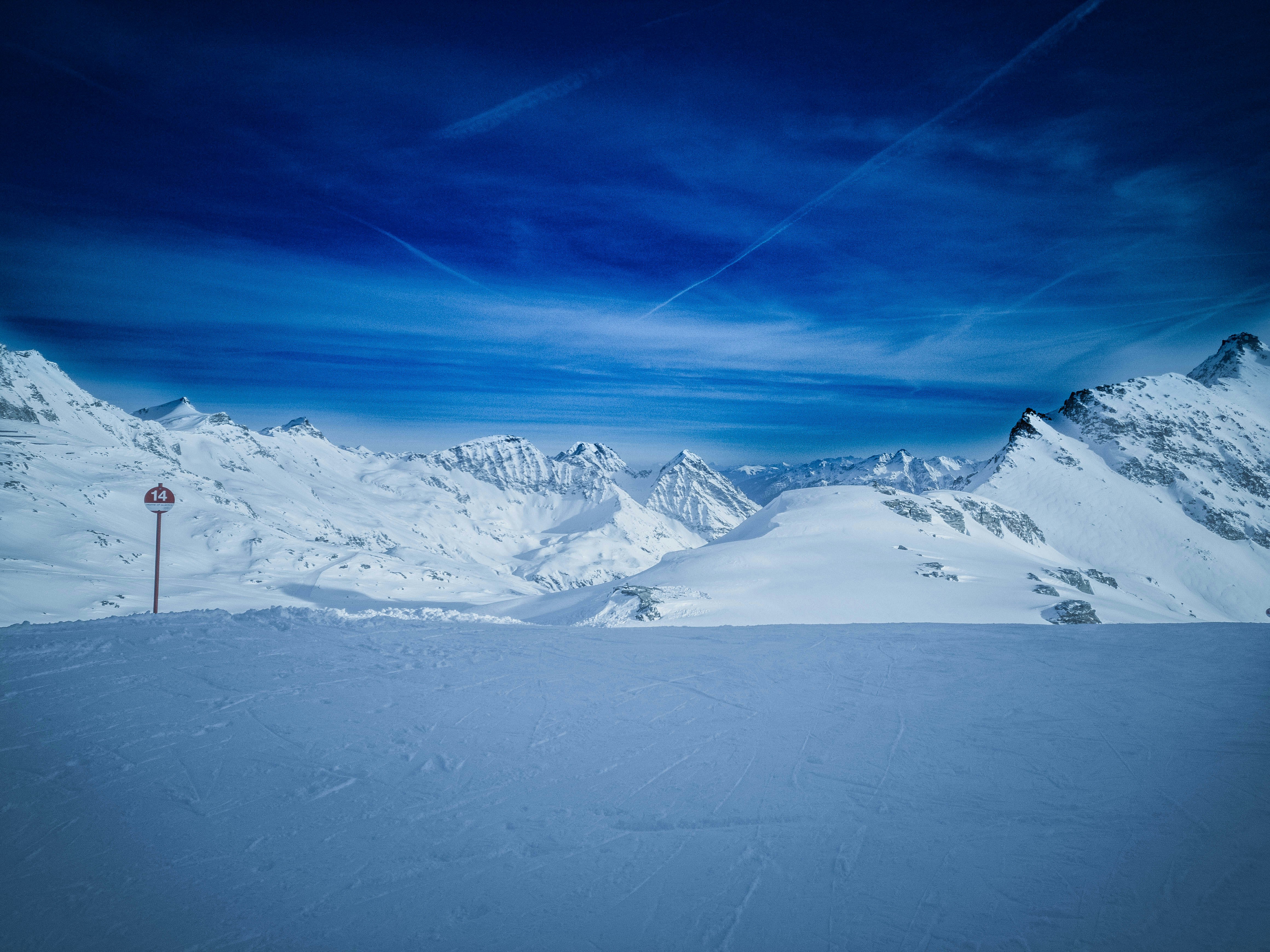 Snow-covered mountain landscape under a vibrant blue sky, featuring distant peaks and a ski trail marker in the foreground.