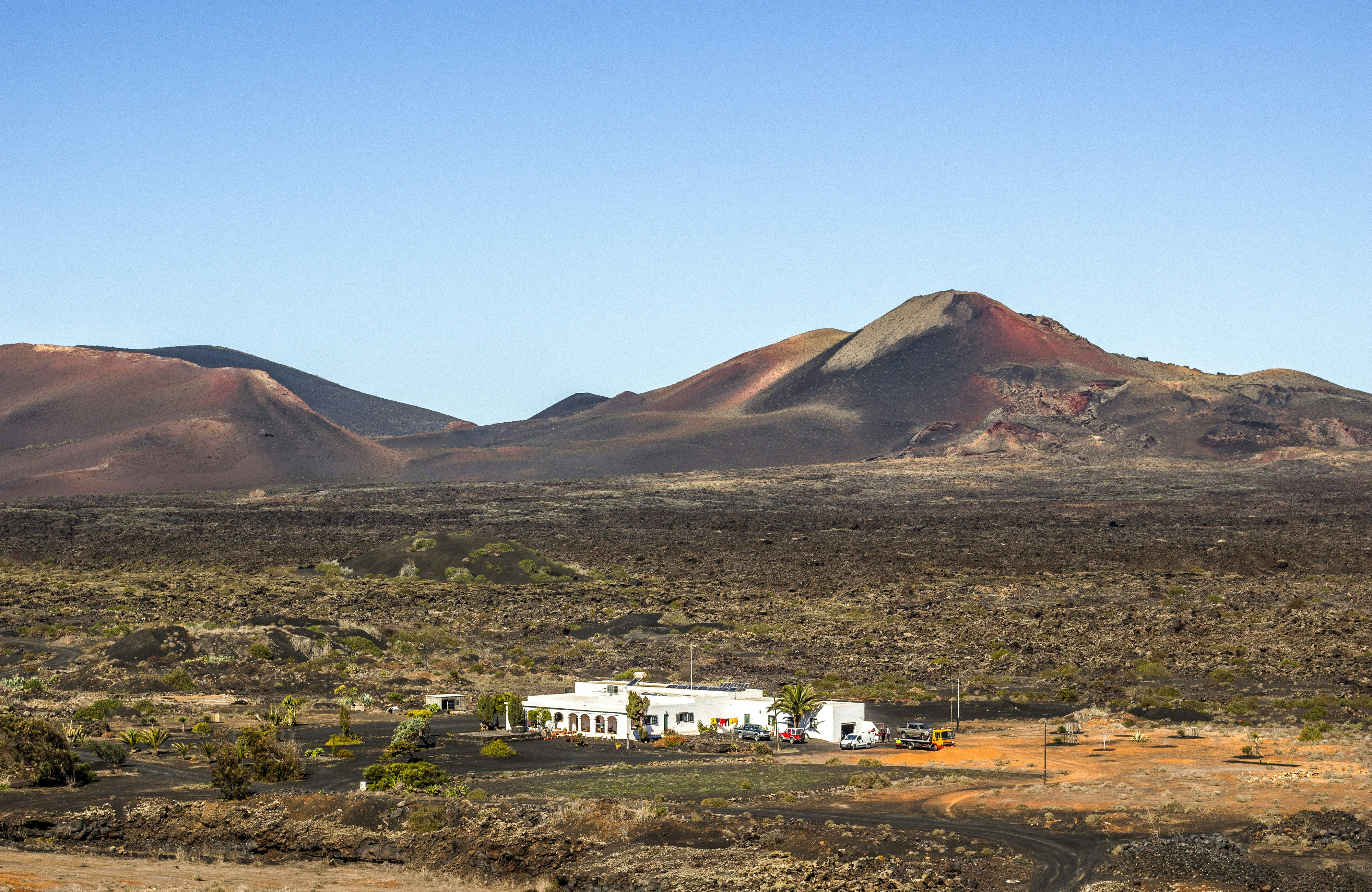 White building complex sits on a barren lava plain with distant multicolored volcanic hills under a clear blue sky.