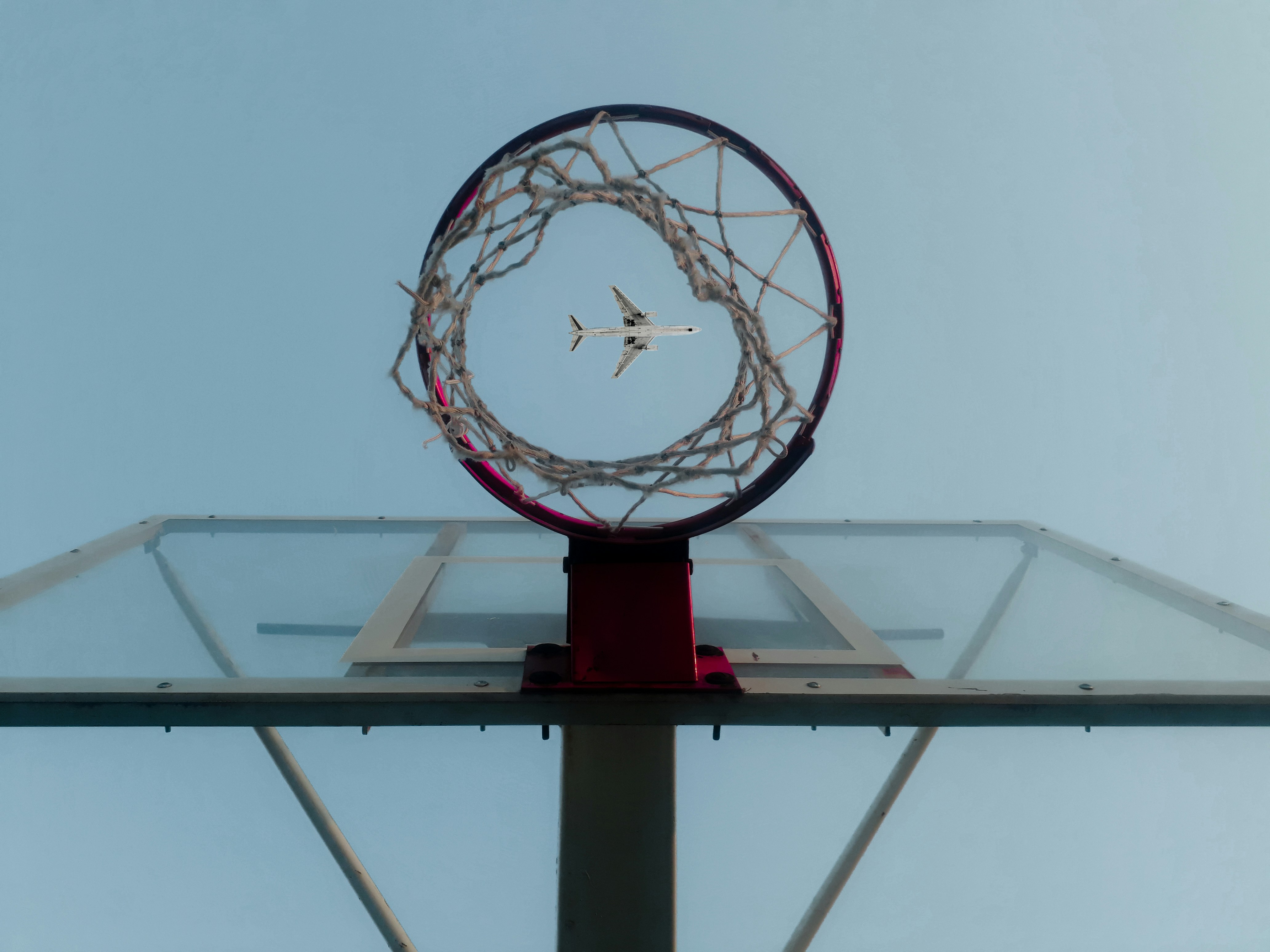 Photograph of a basketball hoop seen from below, with a distant airplane framed in the net against a clear blue sky.