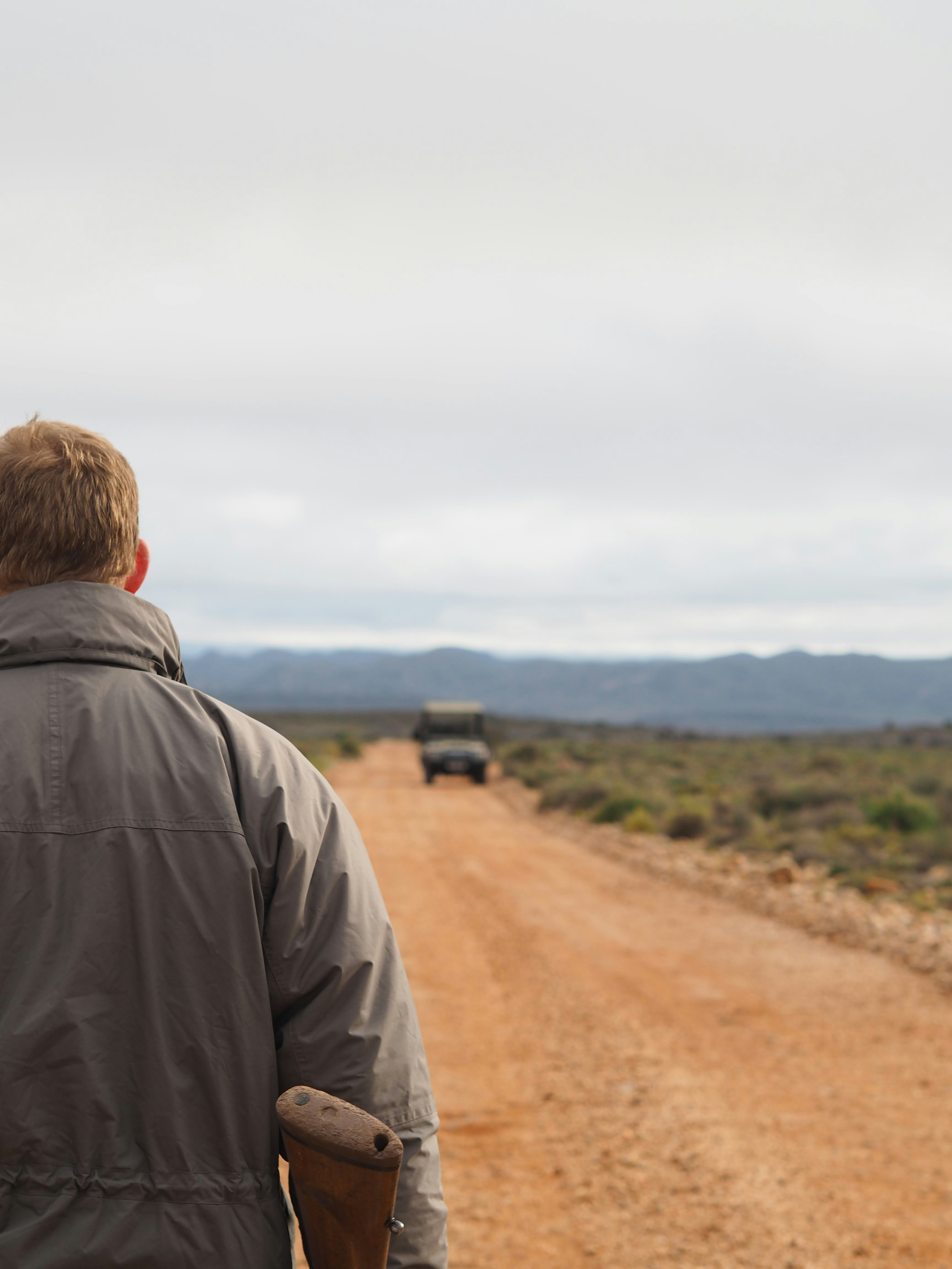 A man with a gray jacket stands at a dusty dirt road, back to the camera, distant jeep on the horizon under a cloudy sky.