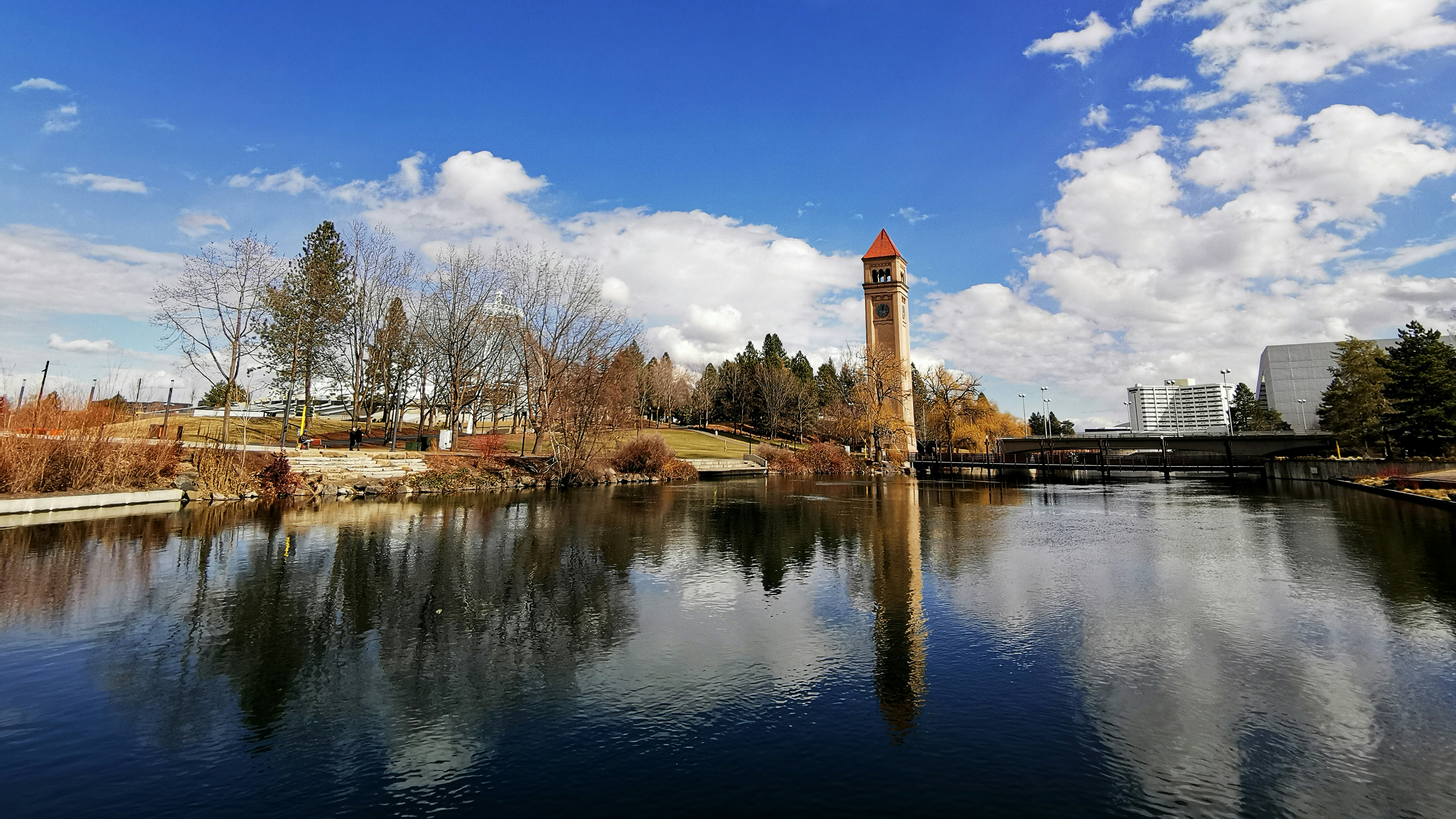 Riverside park with a prominent tower reflecting in calm waters under a bright blue sky.