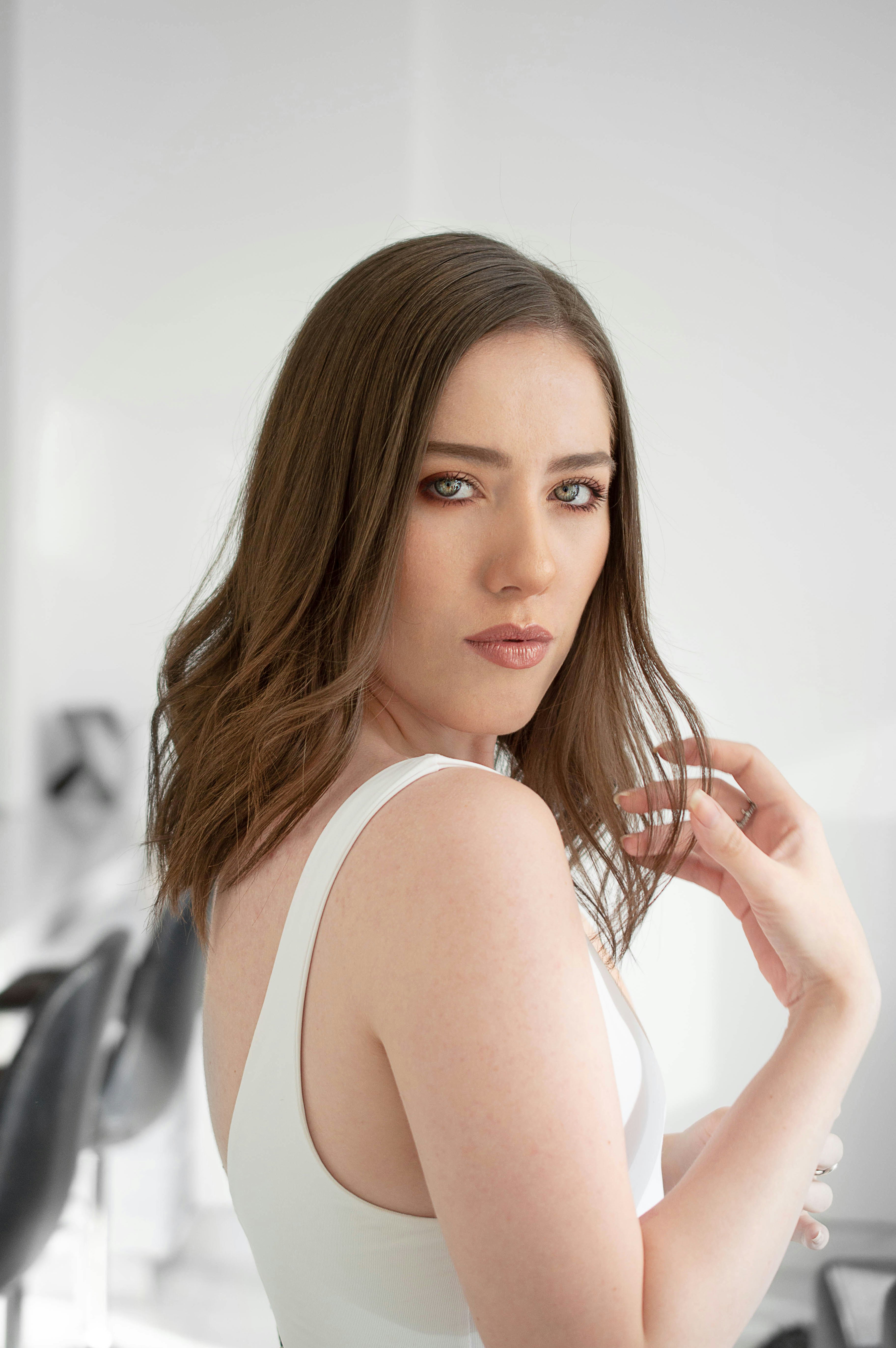 A woman with flowing brown hair gazes over her shoulder, showcasing a sleek white outfit in a bright, modern space.