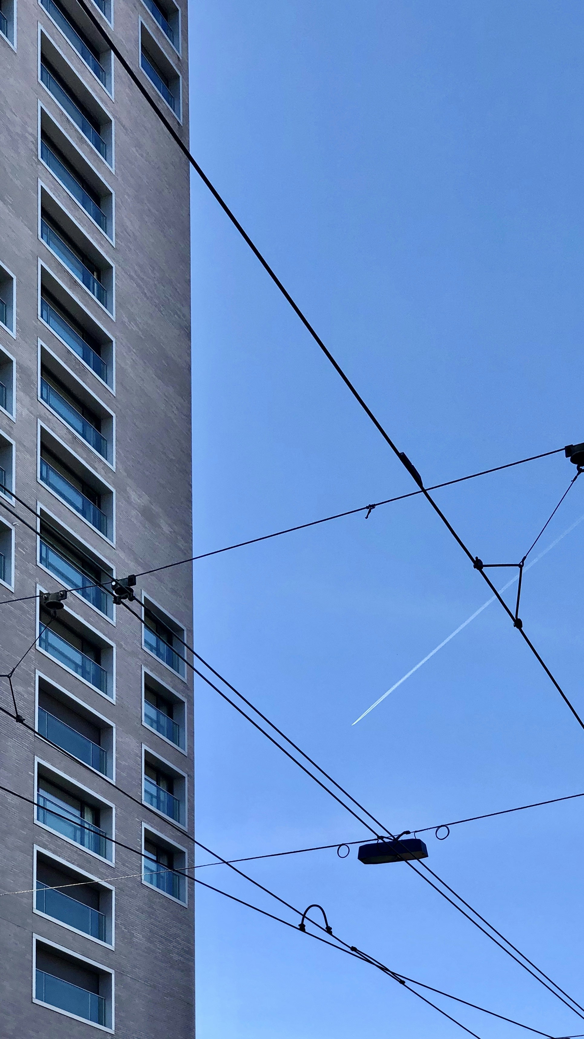 Modern building against a clear blue sky, intersected by overhead wires and a contrail. The composition highlights the interplay of architecture and urban infrastructure.