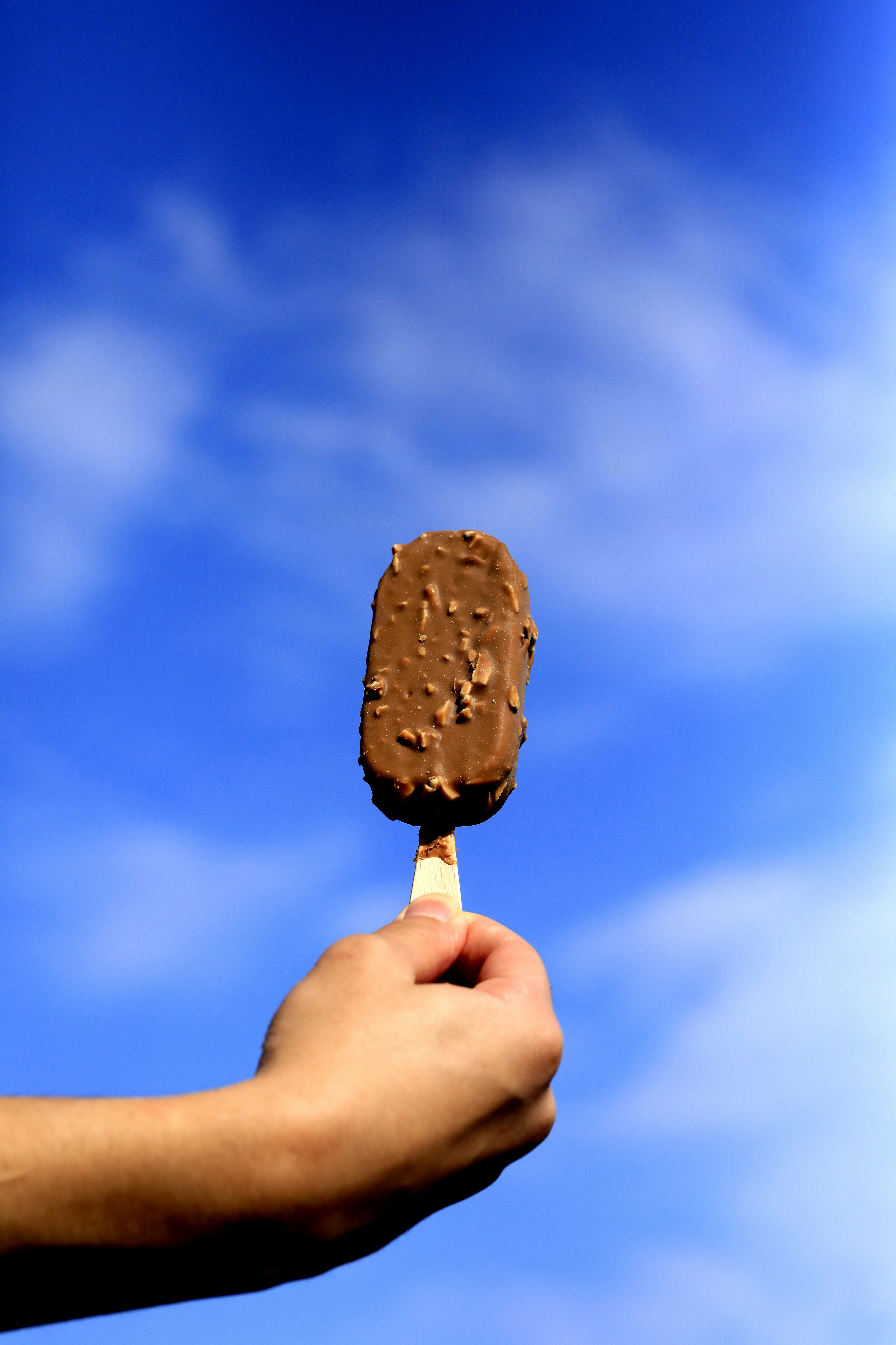 Hand holding a chocolate-covered ice cream bar against a bright blue sky with fluffy clouds.