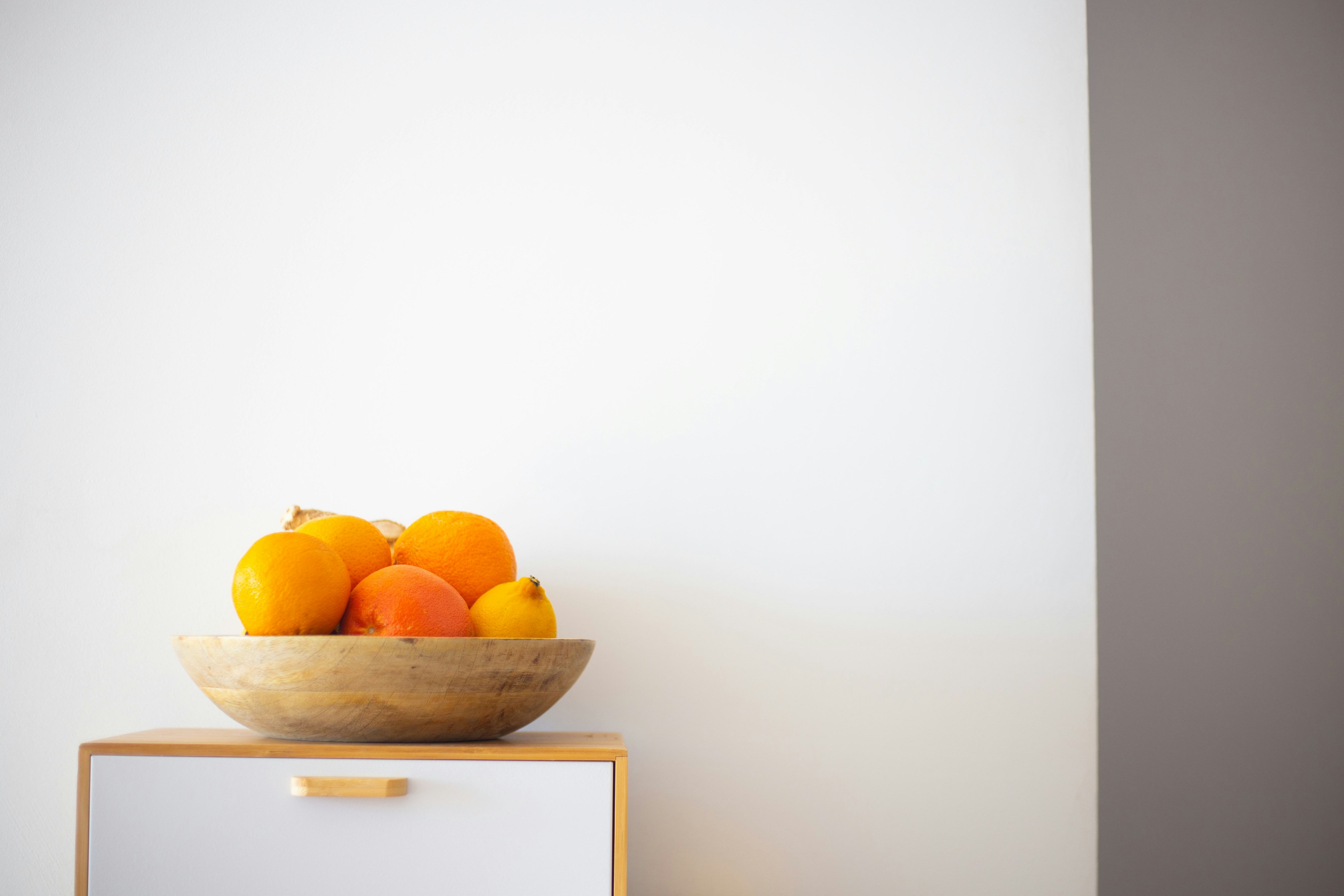 A wooden bowl filled with vibrant oranges and a pear sits atop a minimalist cabinet against a clean white wall.