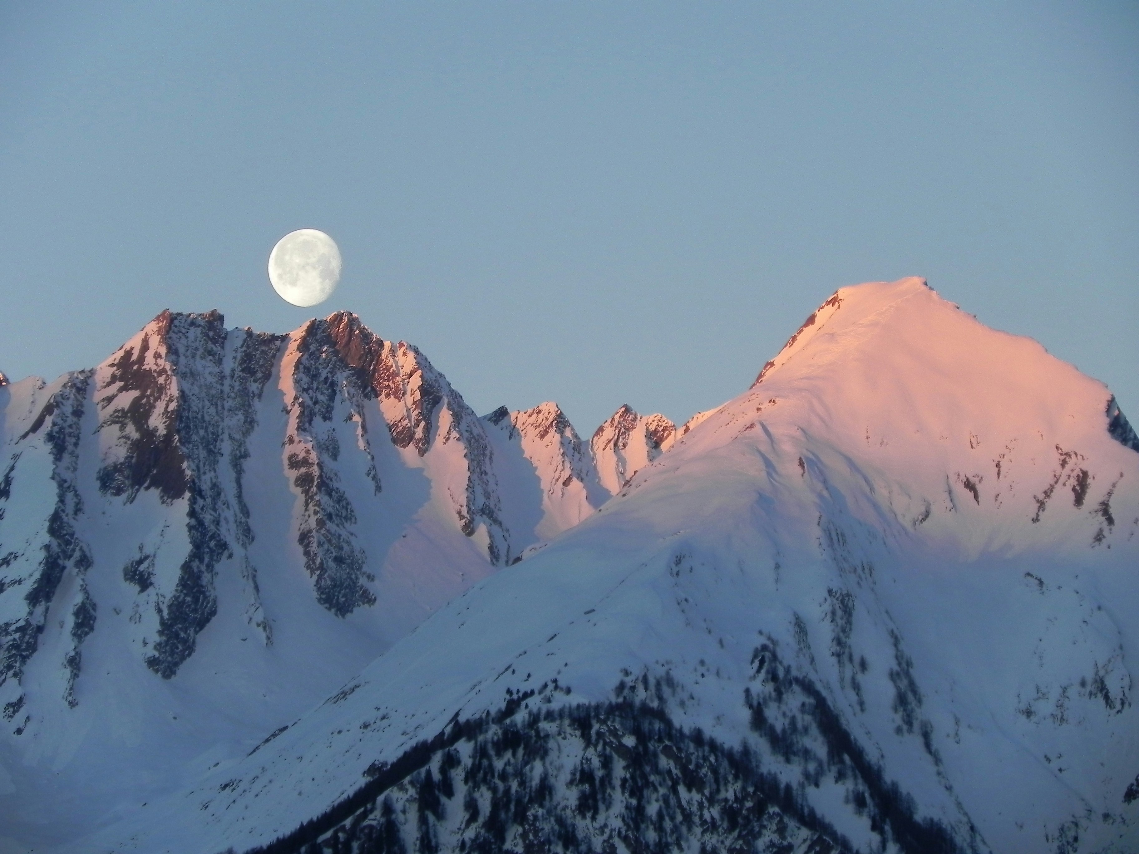 Full moon hovering above snow-capped mountains with a pink sunrise glow.