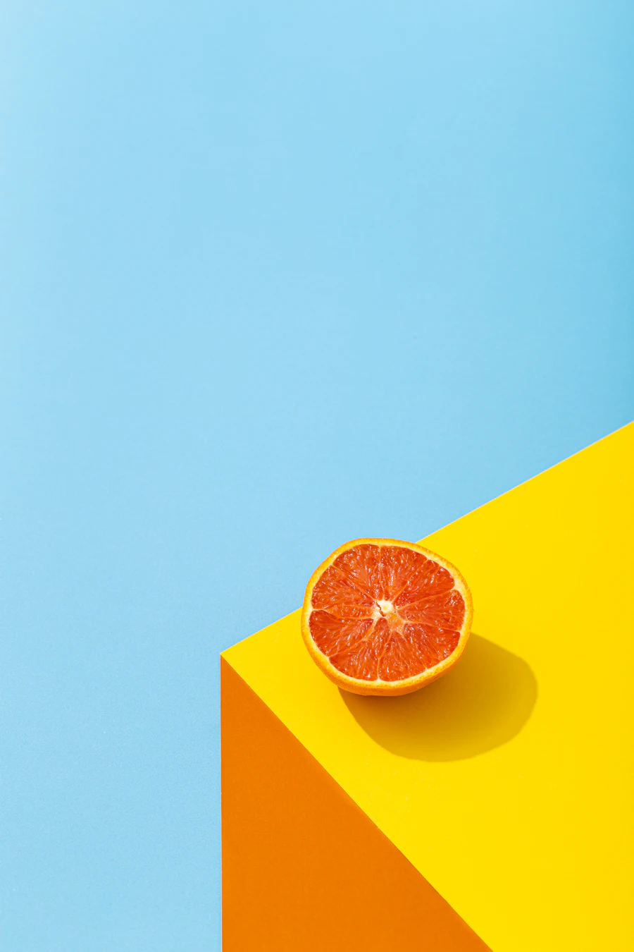 A halved grapefruit rests on a bright yellow geometric block against a soft blue background, with a clean shadow falling to the right.