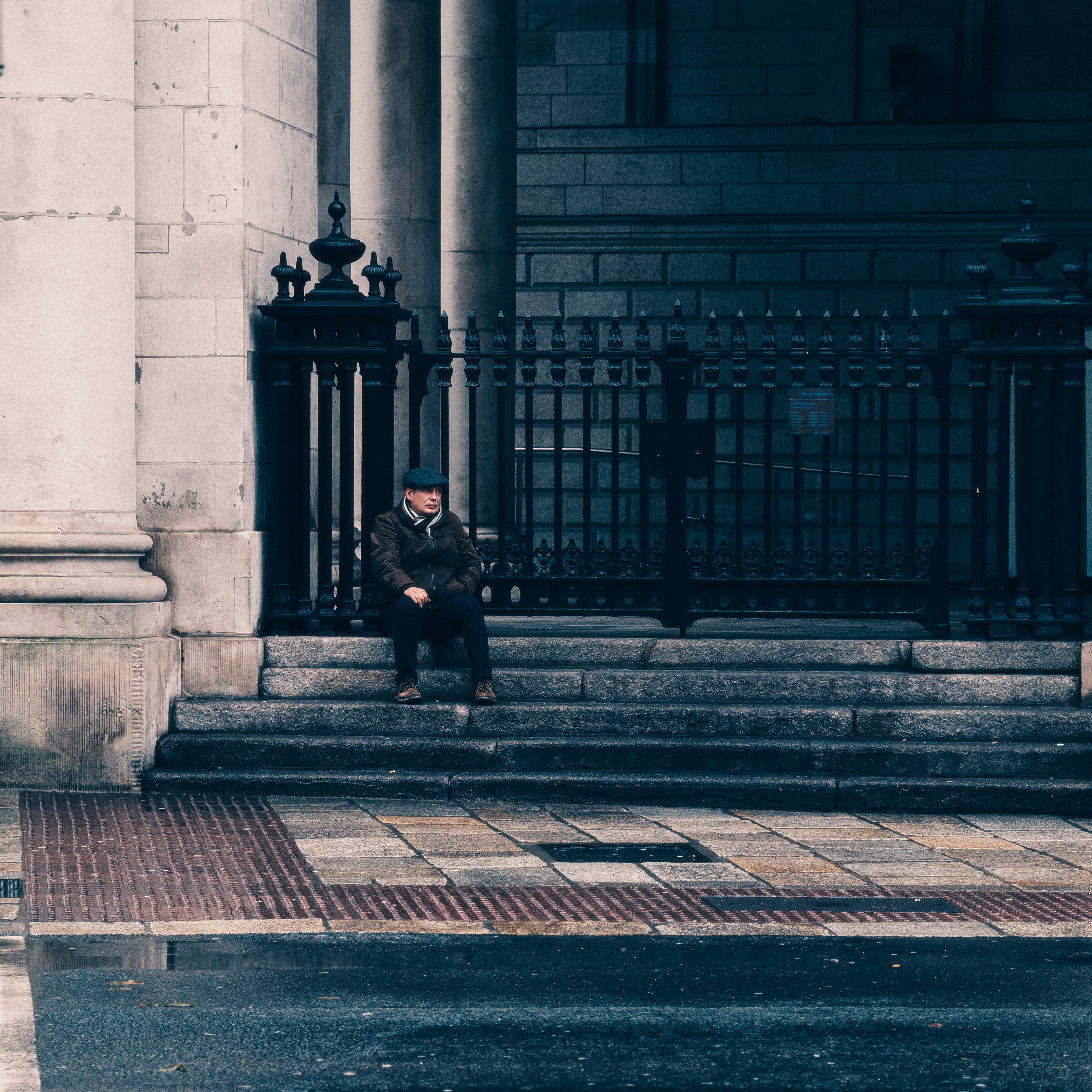 A solitary figure sits on stone steps beneath a grand architectural structure, evoking a sense of contemplation amidst the urban landscape.
