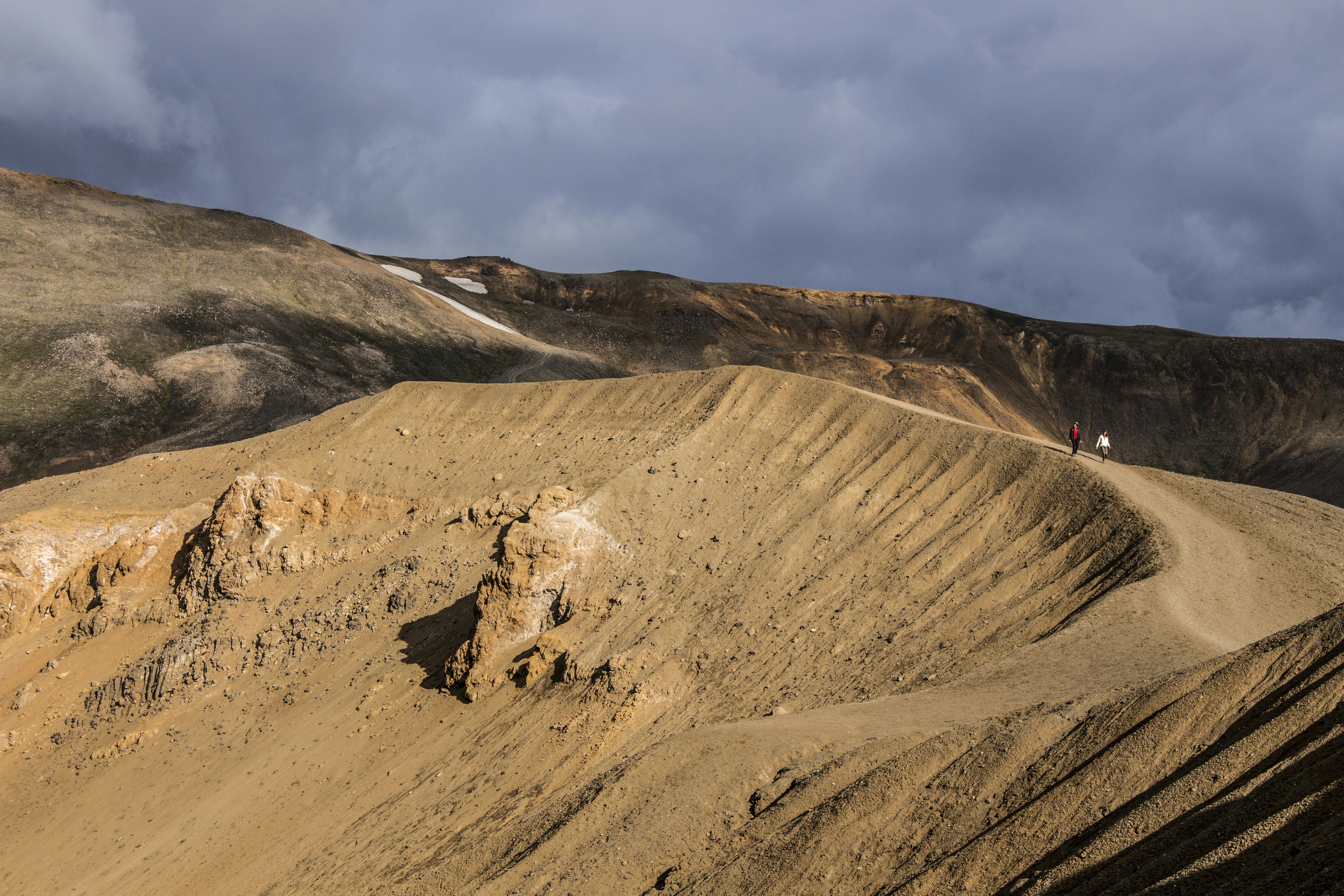 Two people walk along a winding path on a volcanic ridge under a cloudy sky.