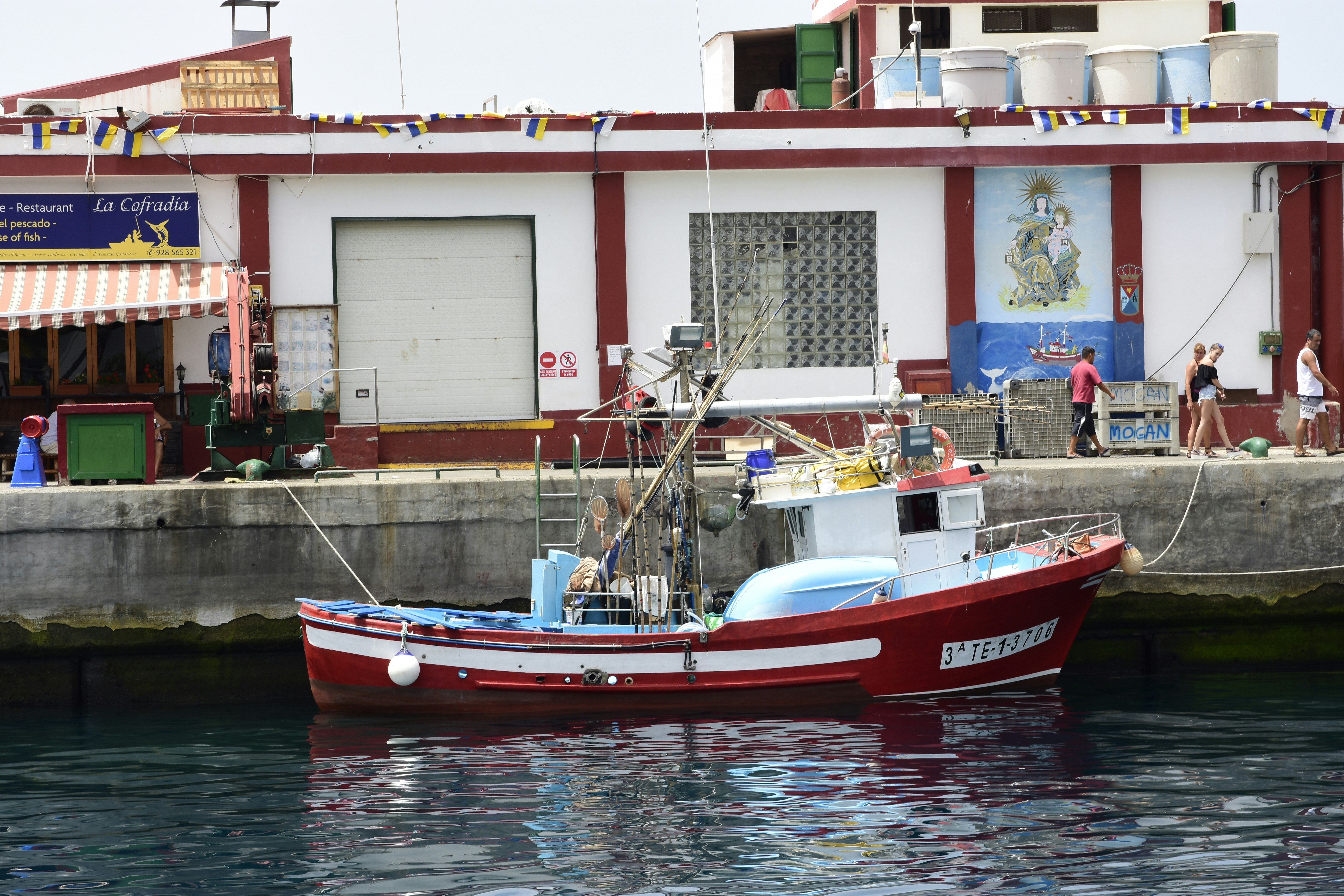 Red and white fishing boat moored at a busy dock with an industrial building in the background.