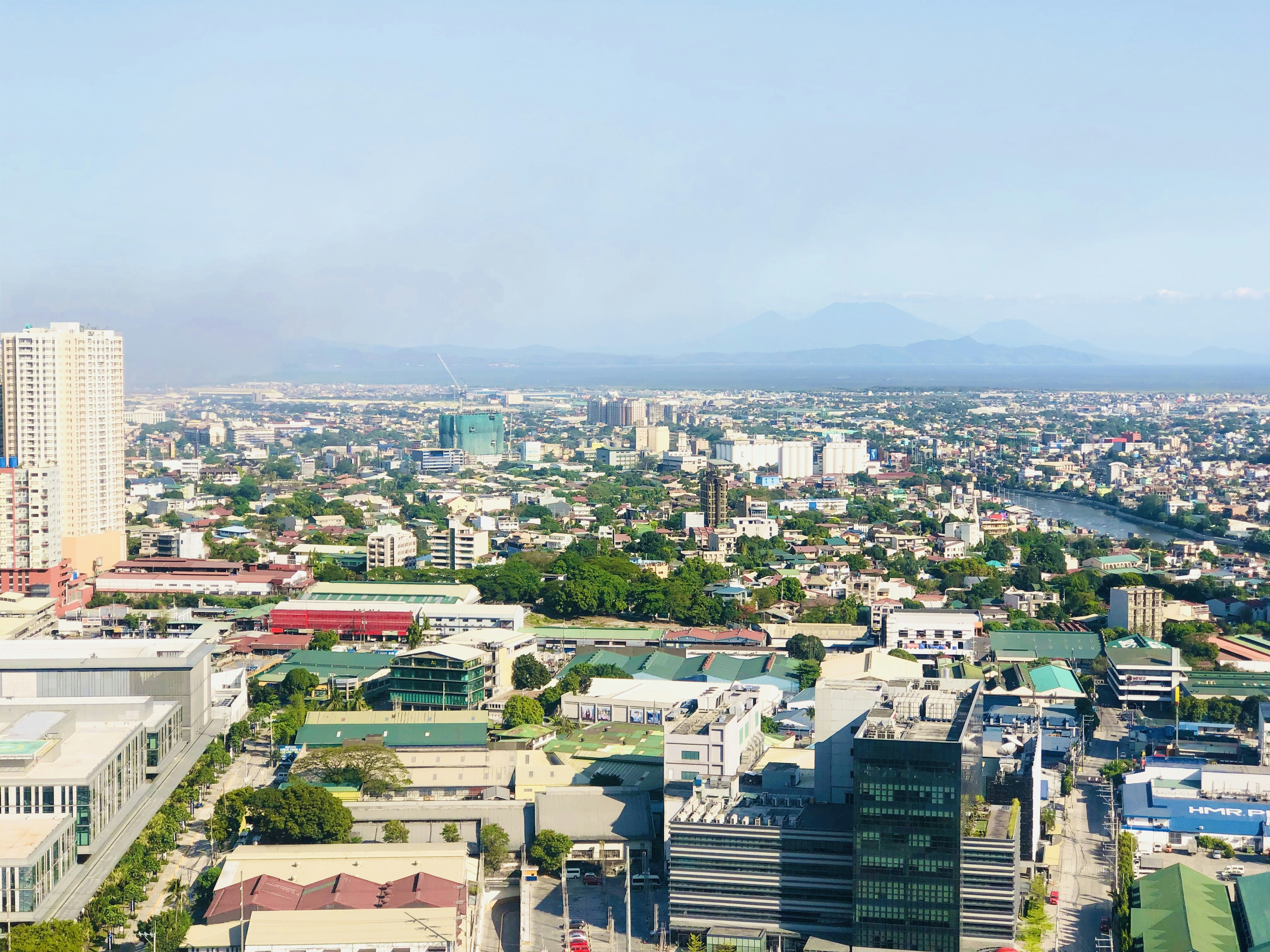A panoramic view of a bustling cityscape, showcasing a blend of modern buildings and greenery along the river. The distant mountains provide a serene backdrop.