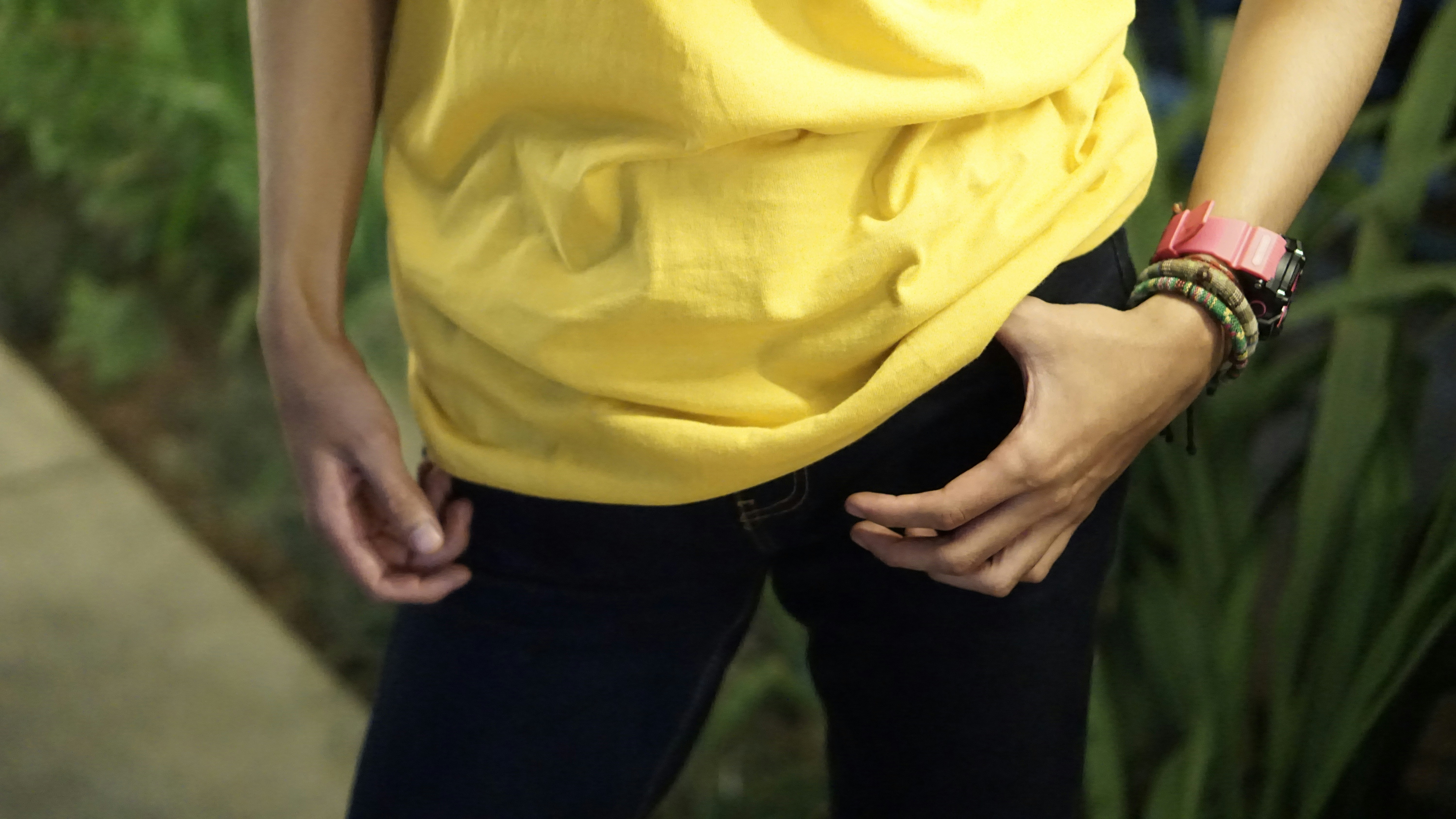A person in a yellow shirt adjusts their jeans while standing amidst lush greenery.