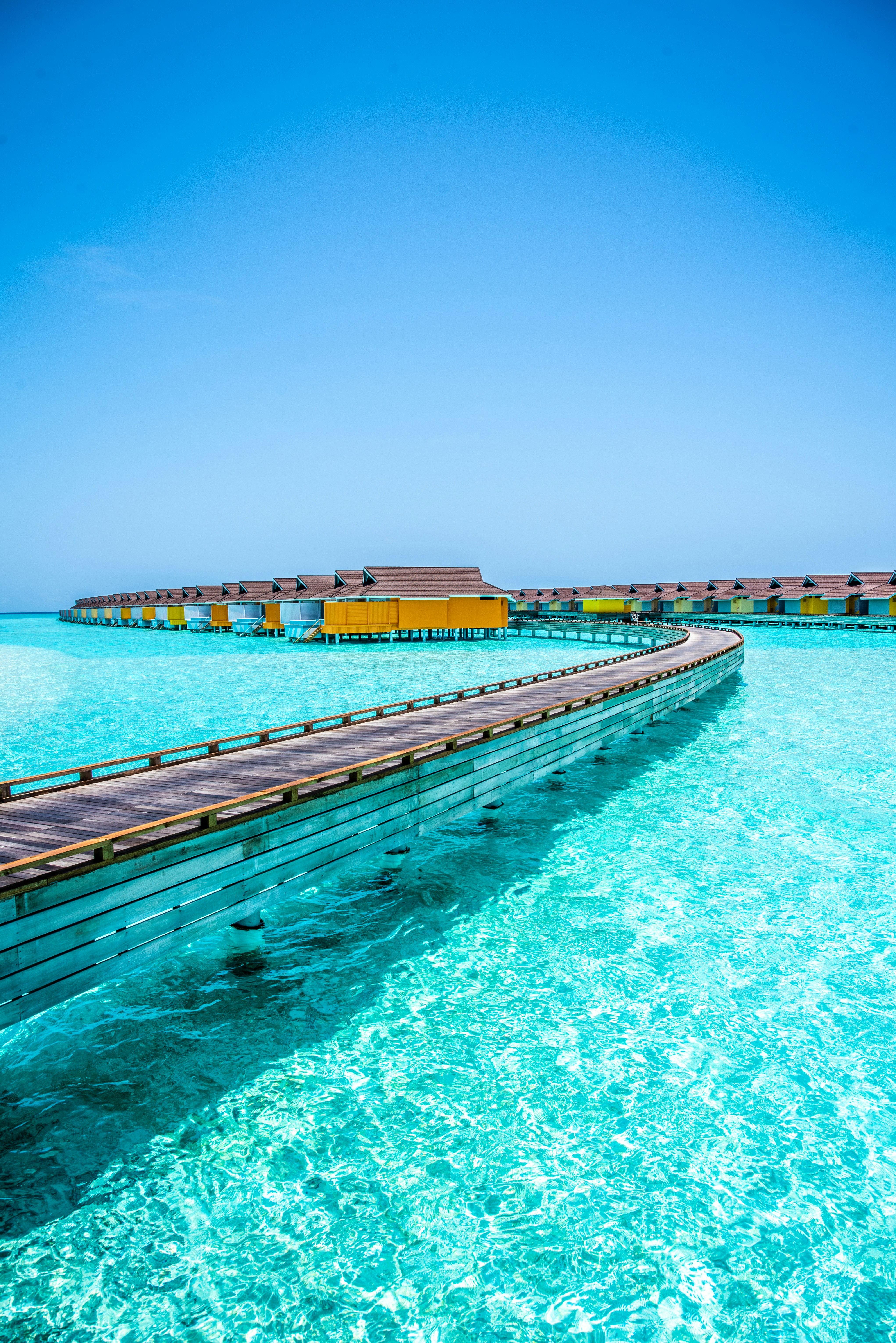 Wooden walkway meandering through crystal-clear waters, leading to overwater bungalows in a tropical paradise.