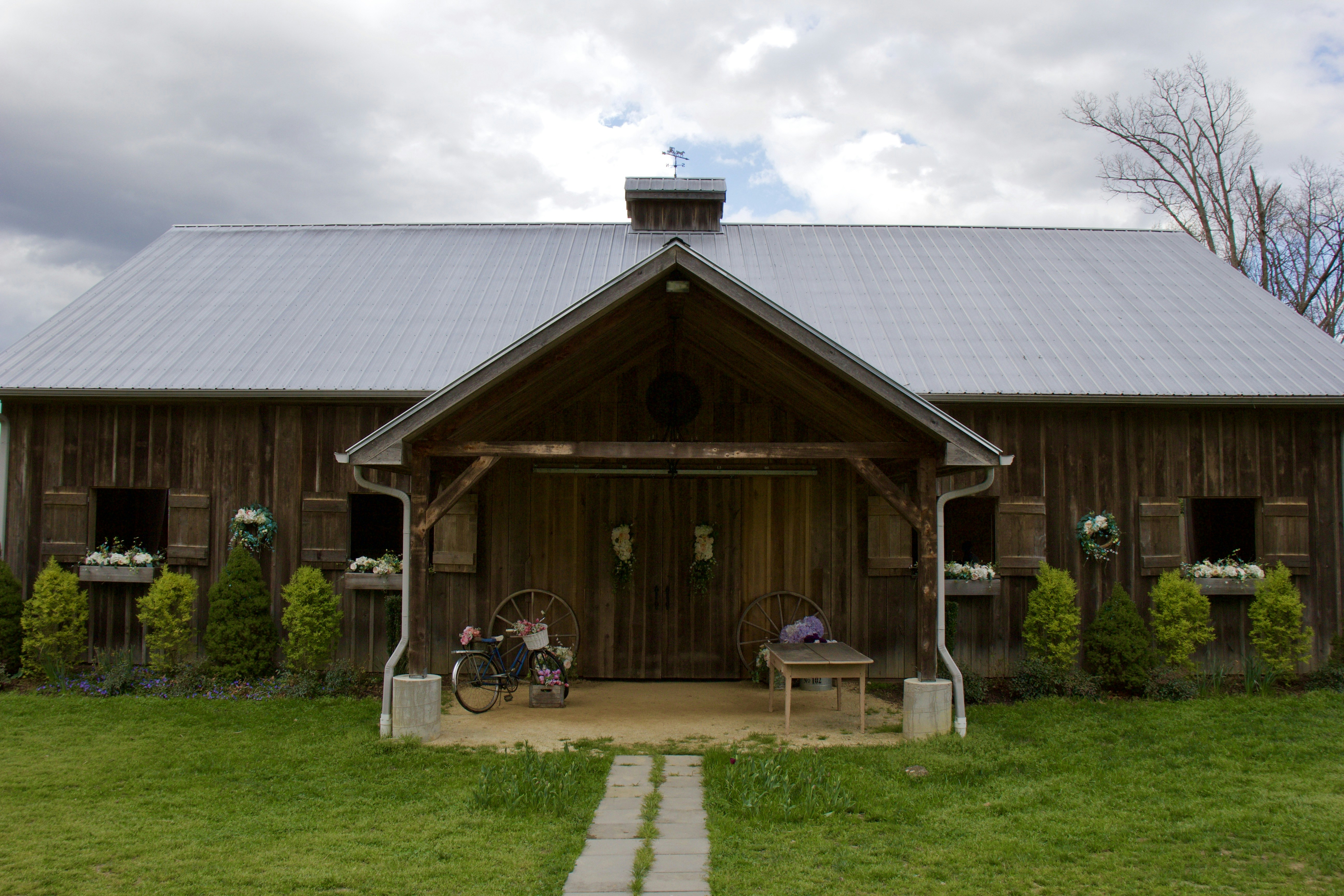 Weathered barn with a welcoming porch adorned with flowers and rustic decor, surrounded by lush greenery.