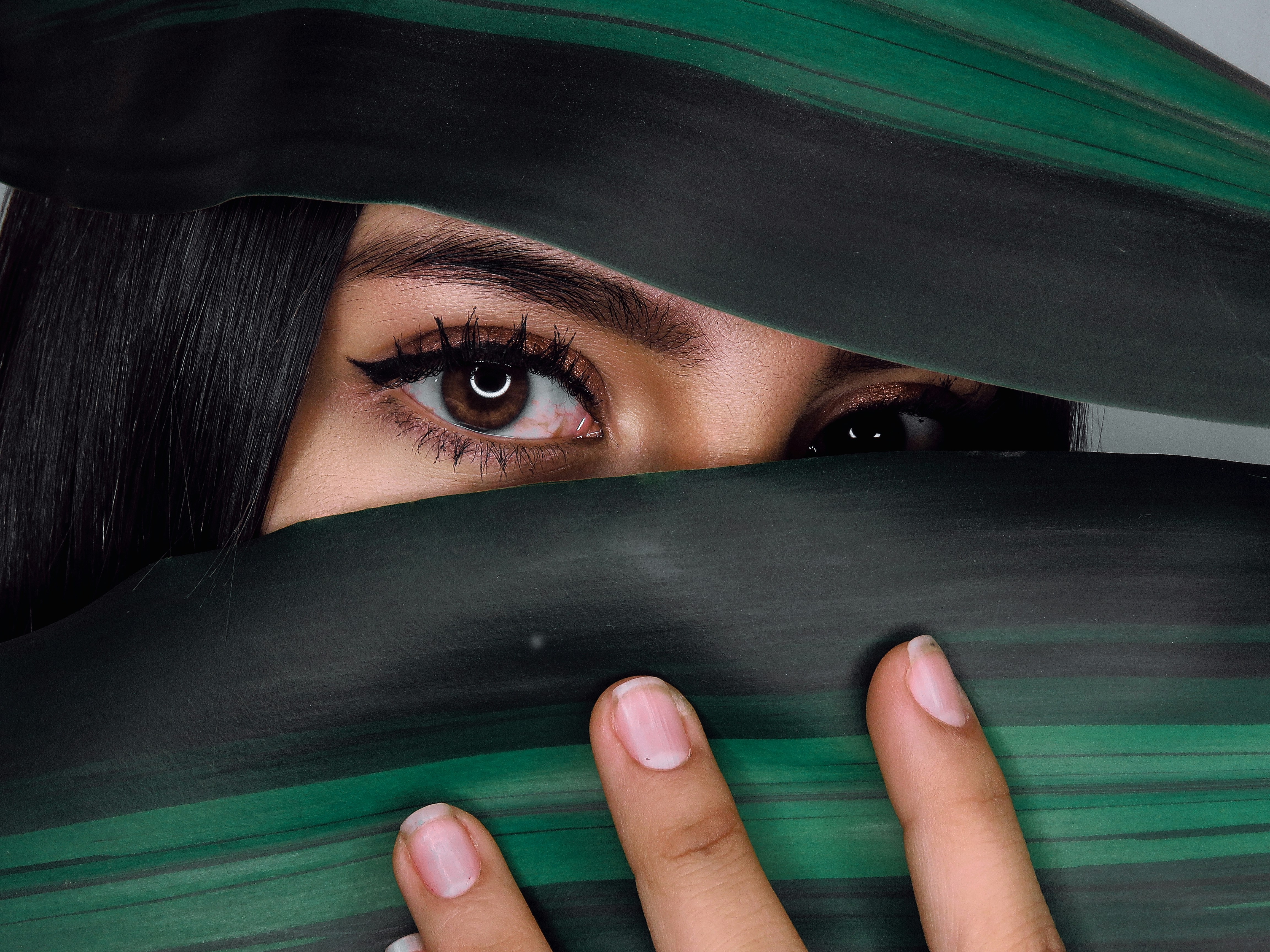 Close-up portrait photograph of a woman peering through layered green leaves with striped bands. The bold eye makeup and partial concealment create a striking focal point.