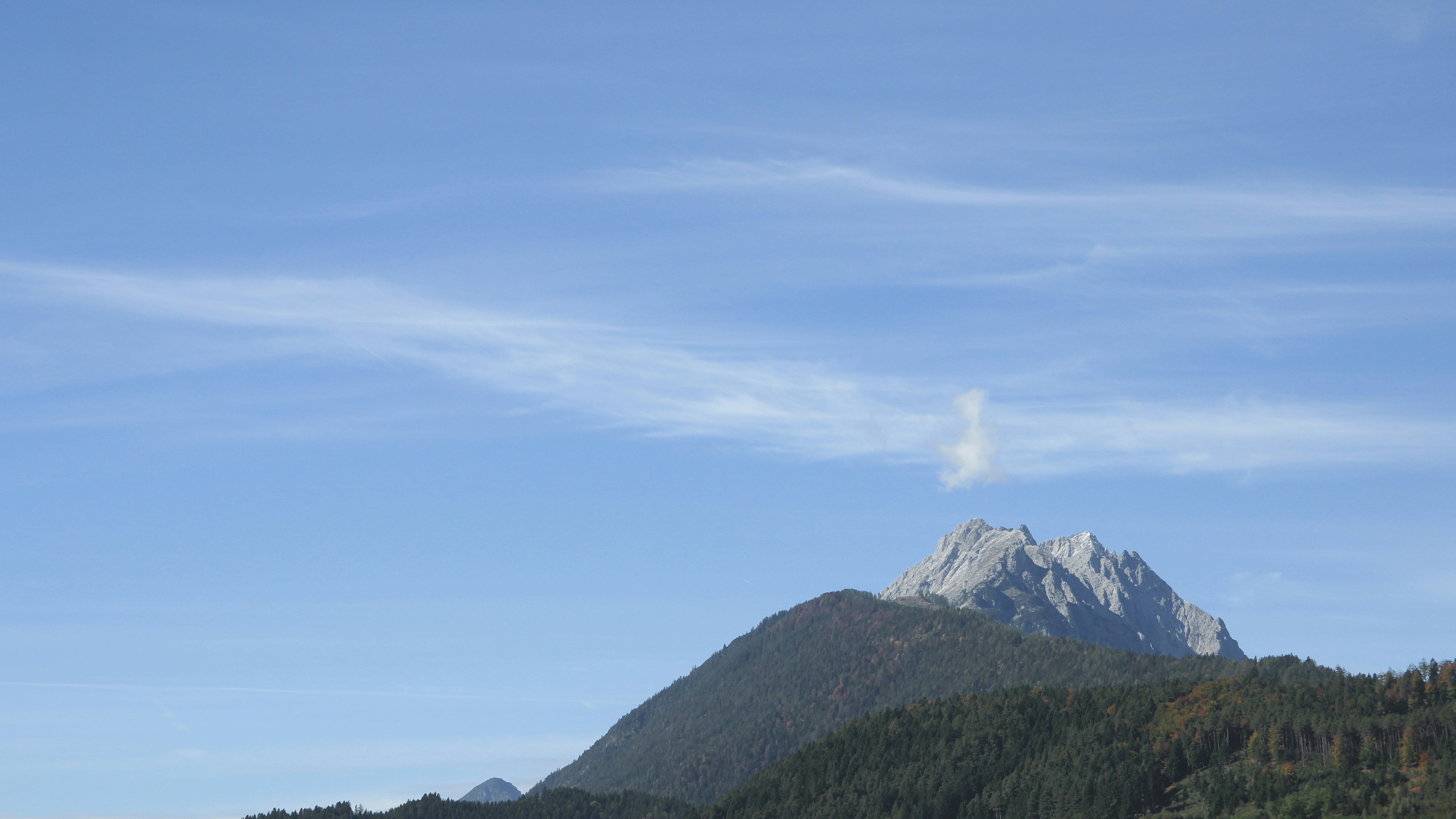 Snow-capped mountain rises above lush green hills under a bright blue sky with wispy clouds.