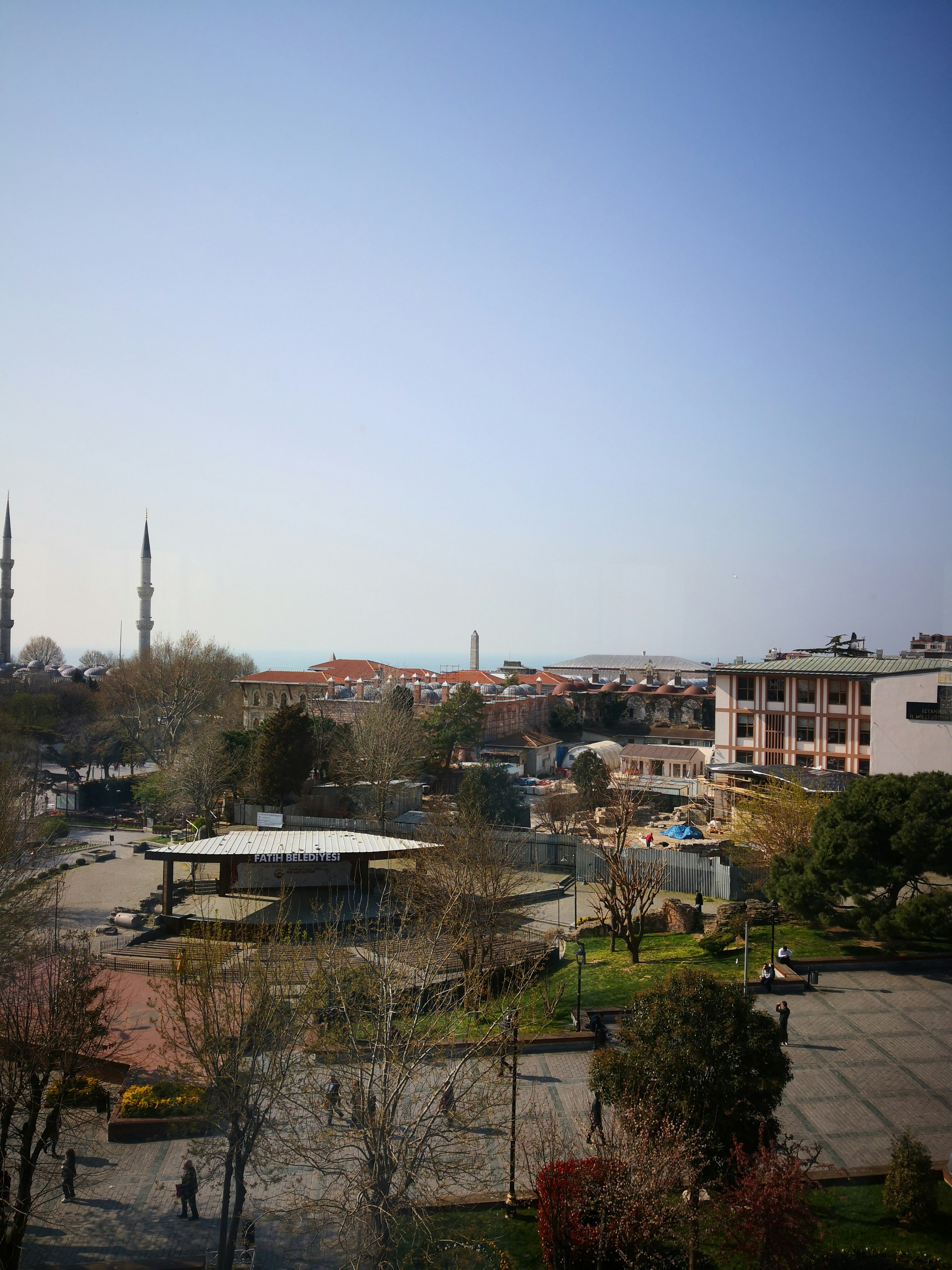 View of a vibrant city square with historical architecture and modern structures, framed by minarets and greenery.