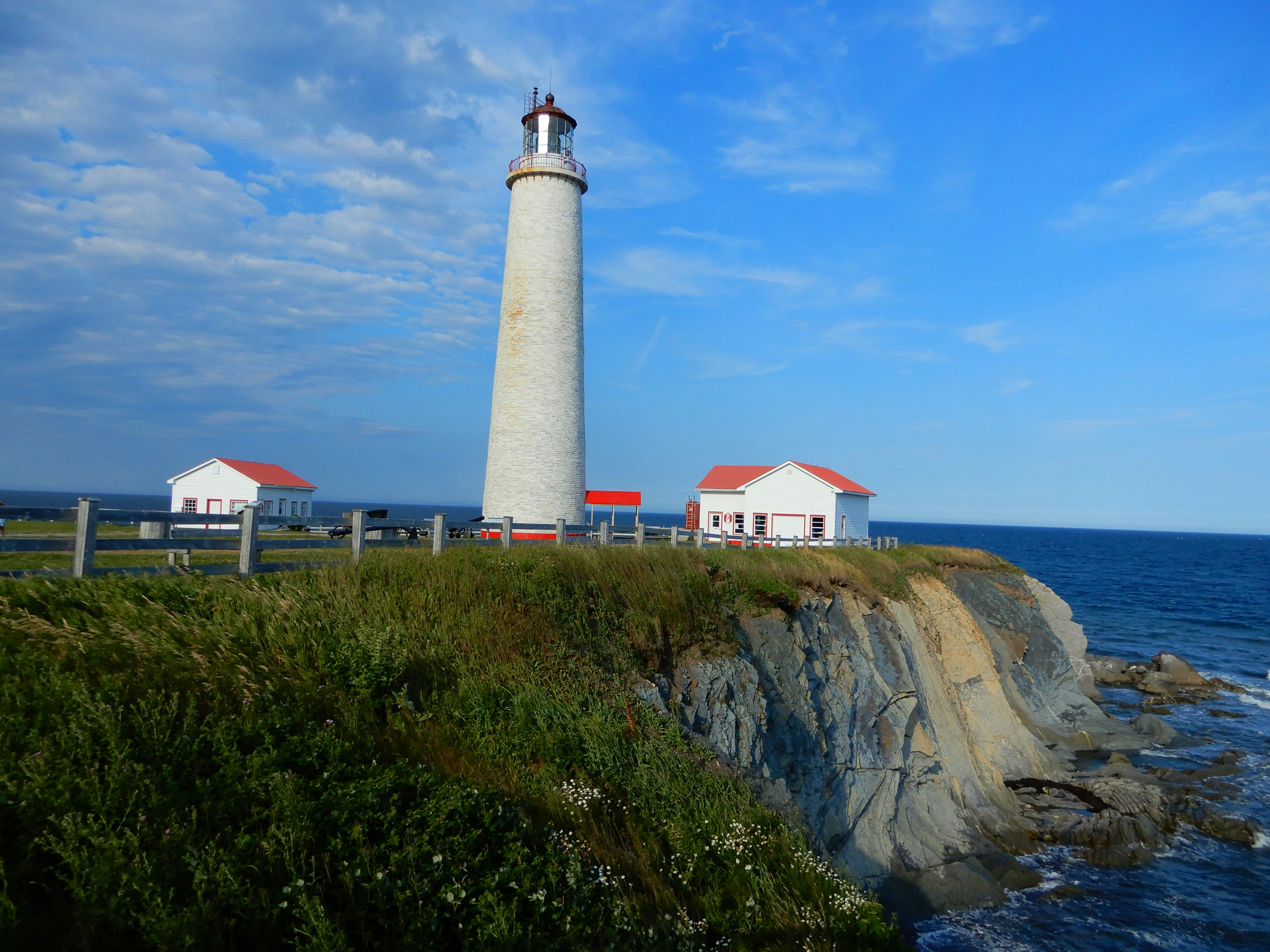 A tall lighthouse stands proudly on a rocky cliff, flanked by charming white buildings under a blue sky with scattered clouds.